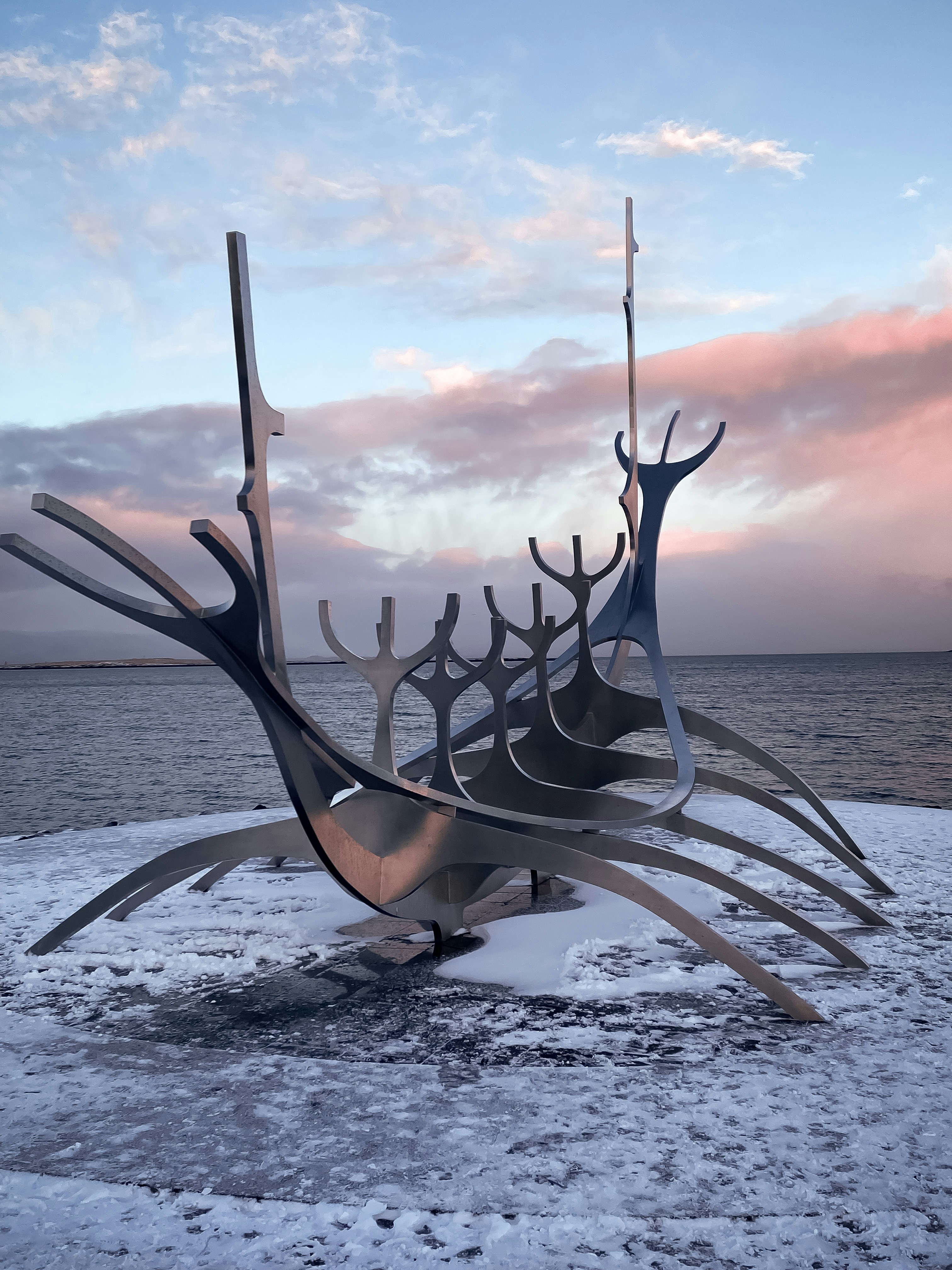 Sleek metal sculpture resembling a ship's bow, set against a snowy landscape and serene sea under a pastel sky.