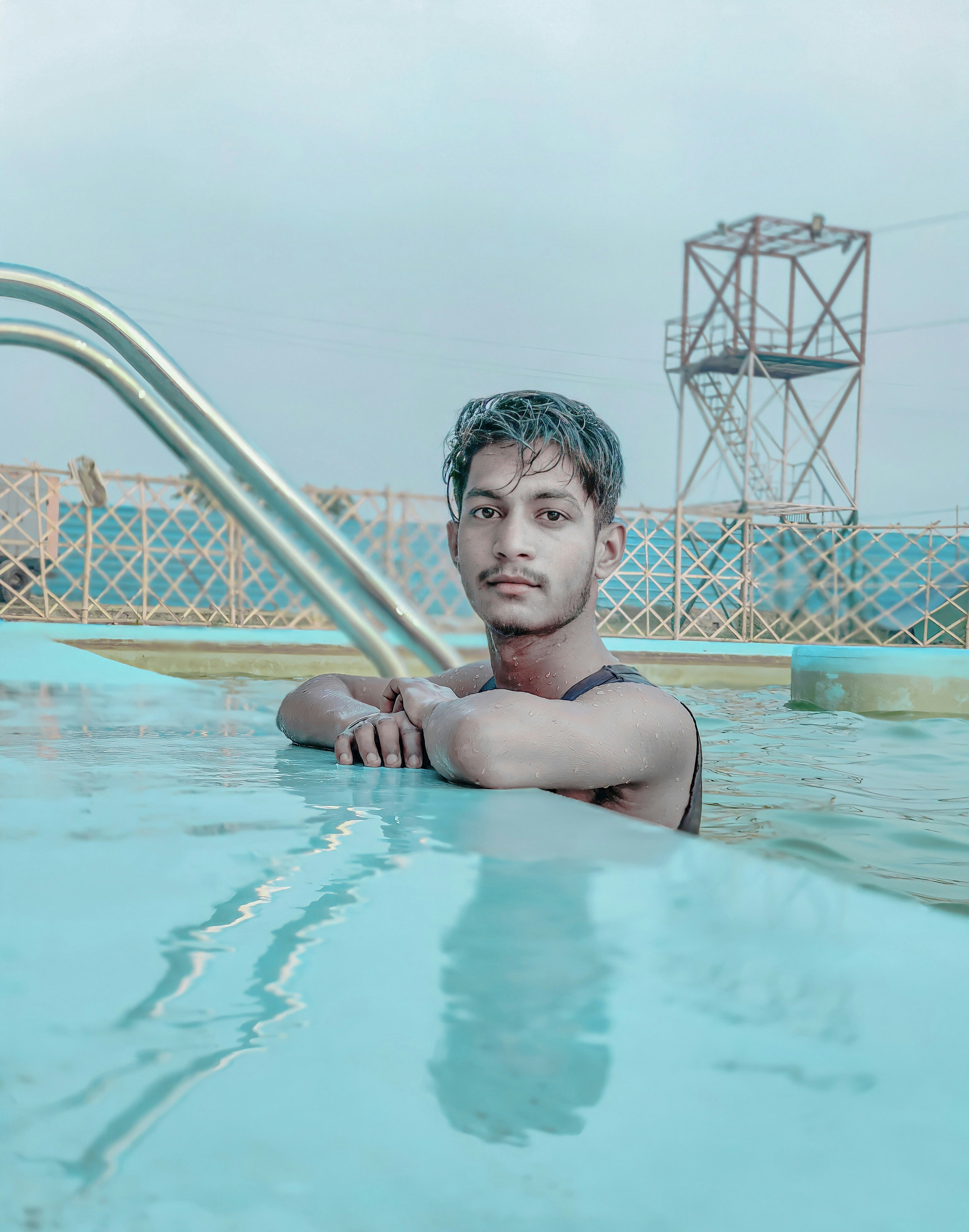 Young man relaxing in a turquoise pool, leaning on the edge with a serene expression. The background features a faint structure and a clear sky.