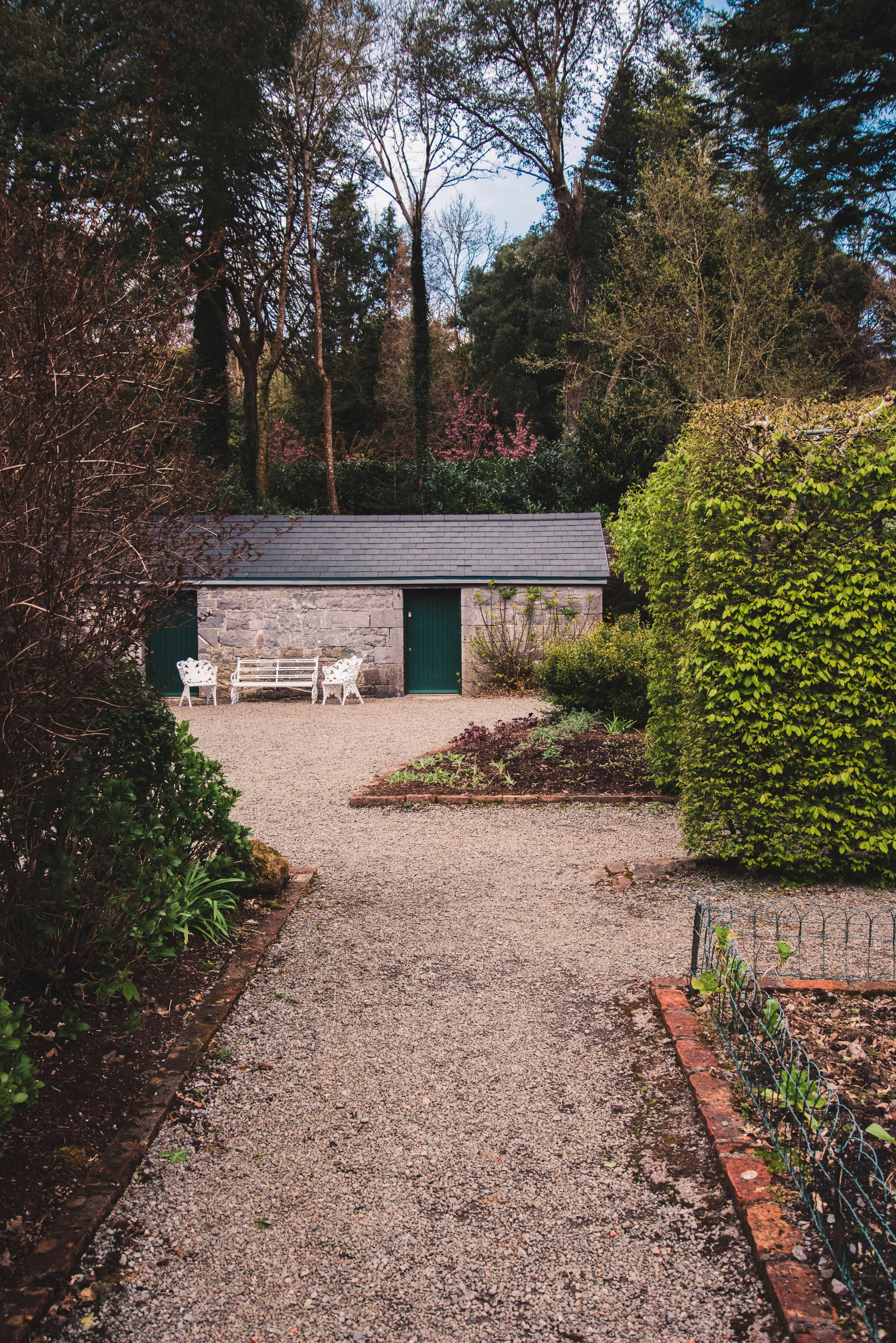 Charming garden pathway leading to a quaint stone shed, framed by lush greenery and blooming trees. A pair of white benches invite relaxation.