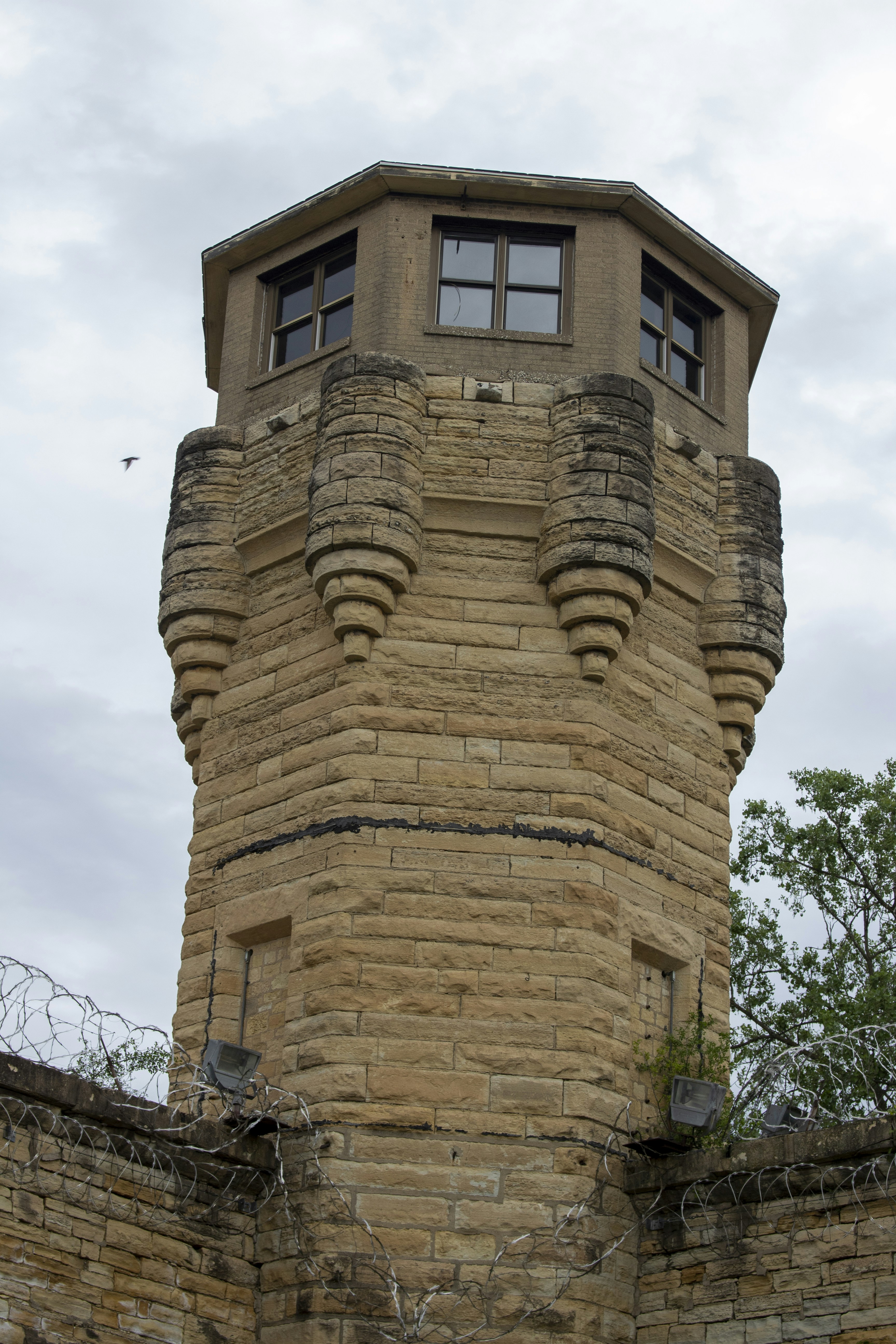 Historic stone watchtower with intricate architectural details, surrounded by barbed wire, under a cloudy sky.