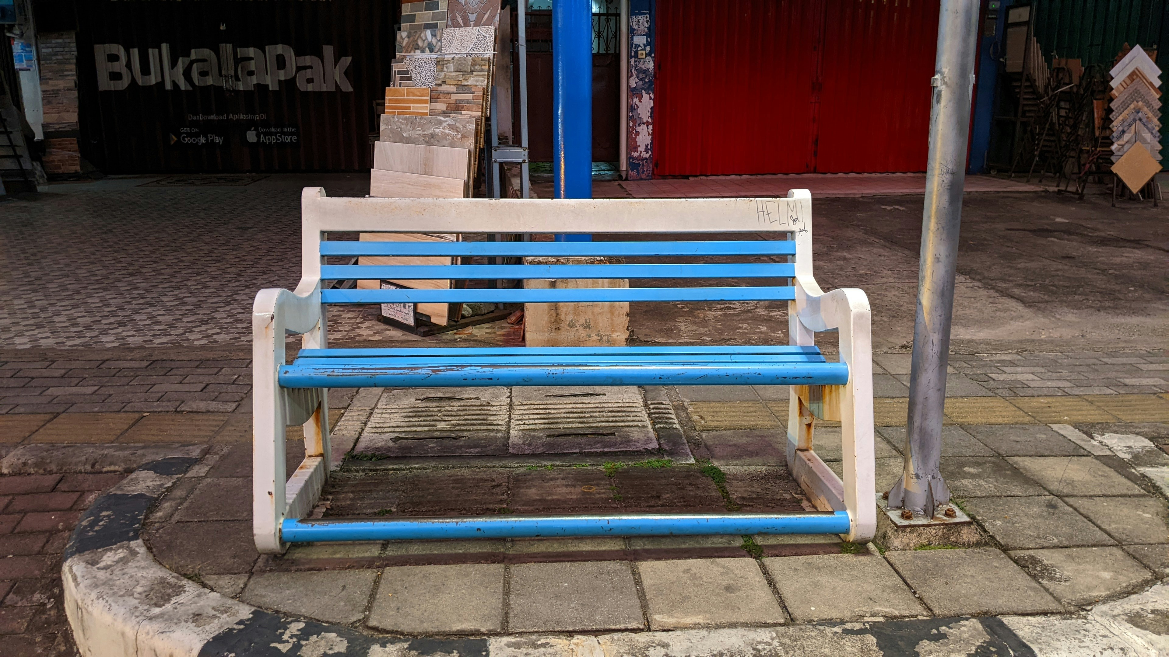 Photograph of a white bench with blue slats on a cobblestone street, storefronts visible in the background.