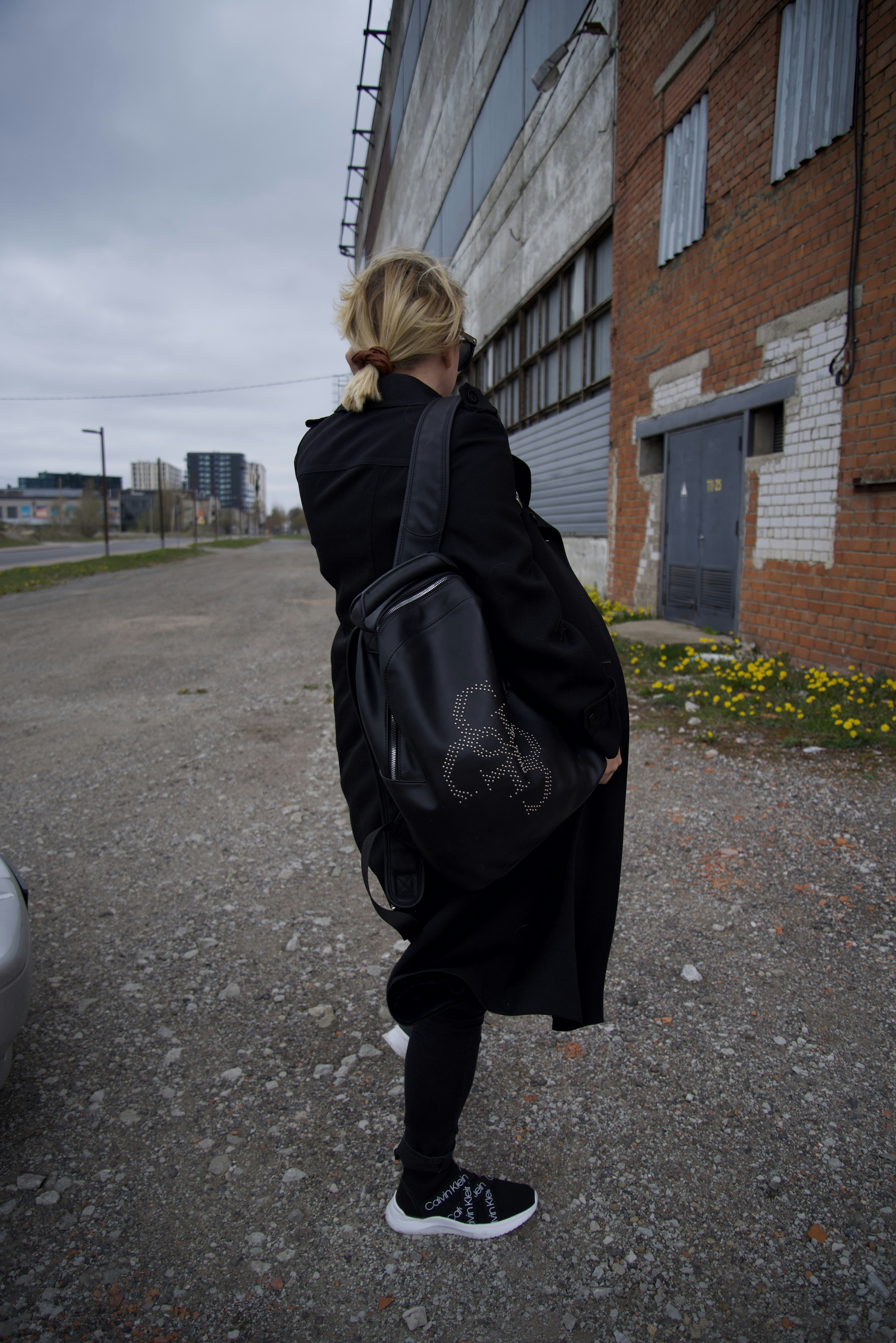 A person in a black coat stands with their back to the camera, gazing at a deserted urban landscape filled with industrial buildings and wildflowers.