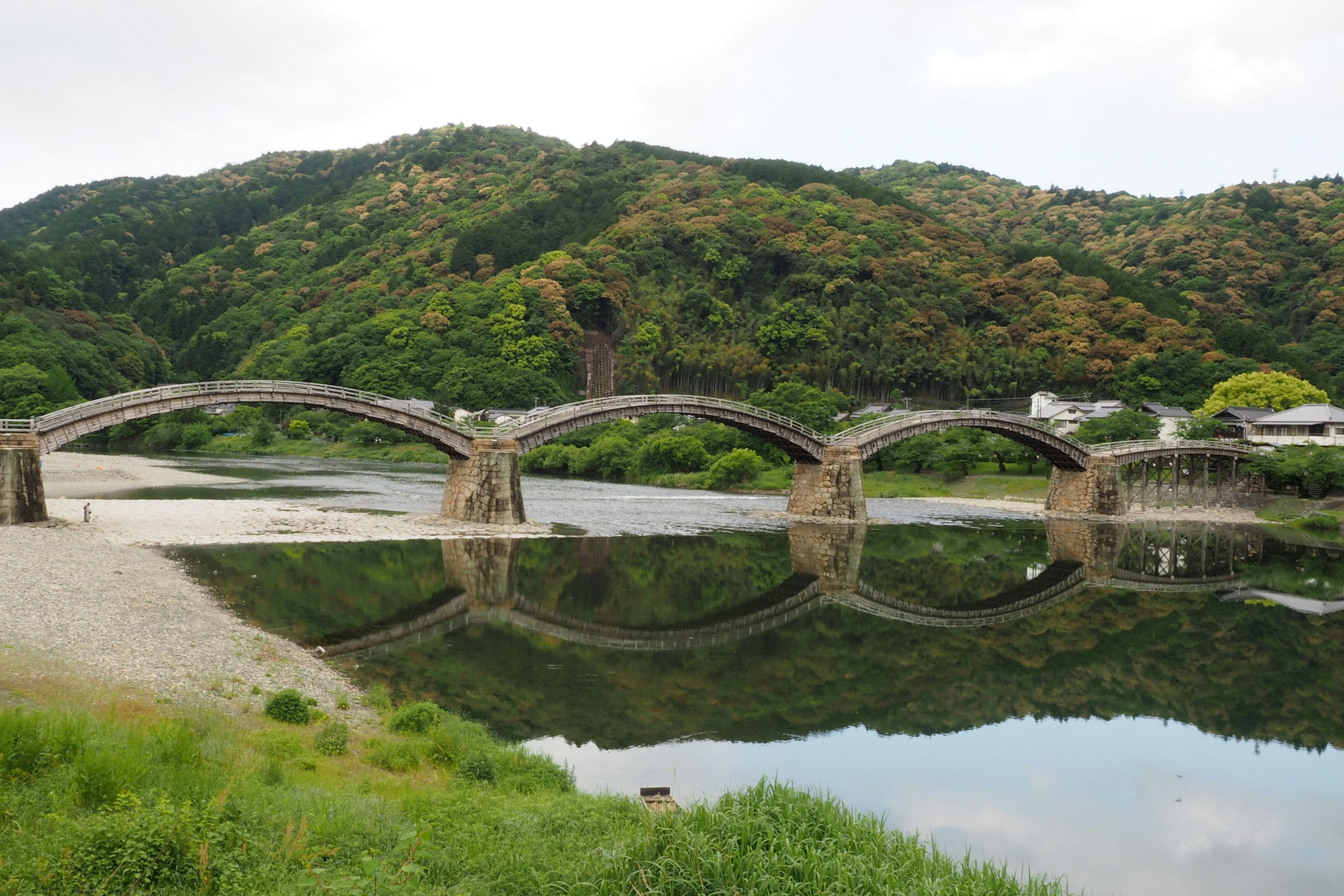 Arched wooden bridge reflecting in a calm river against a backdrop of lush green hills.