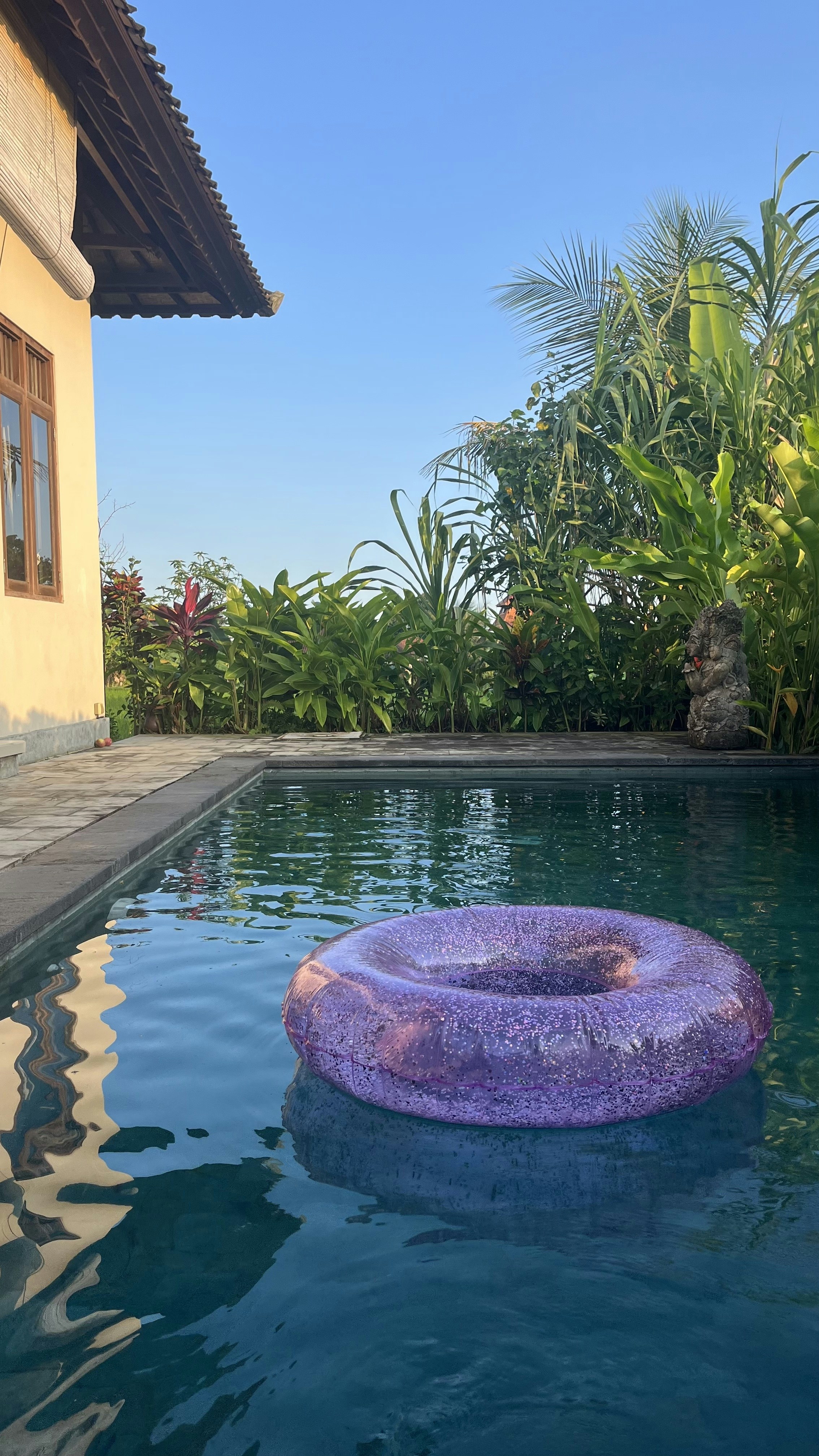 Purple glittery pool float resting on calm water, surrounded by lush greenery and a warm, inviting villa. The clear blue sky enhances the tranquil atmosphere.