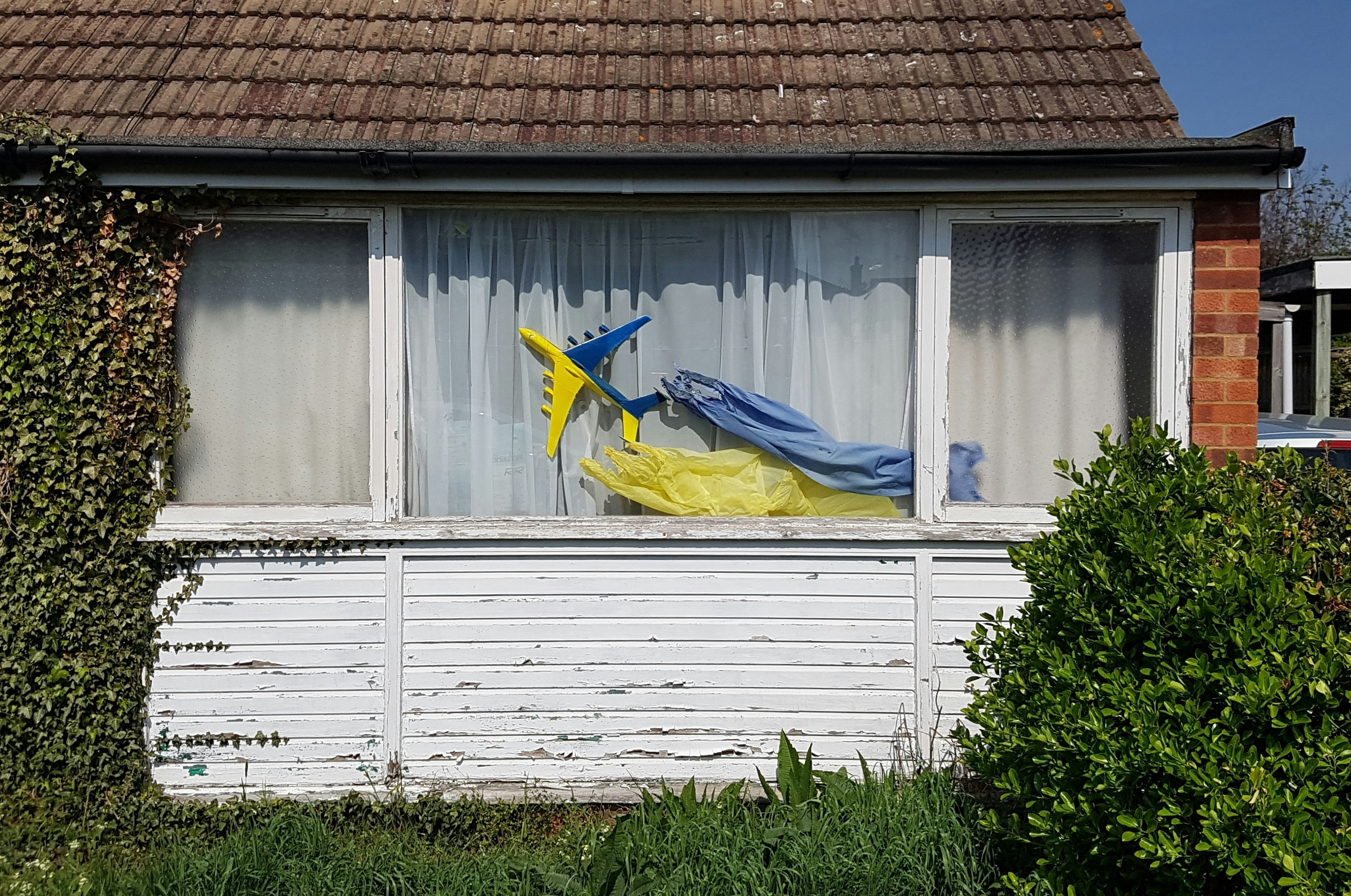 A vibrant yellow and blue model airplane rests on a hand-shaped decoration in a sunlit window, framed by lush greenery and a rustic house facade.