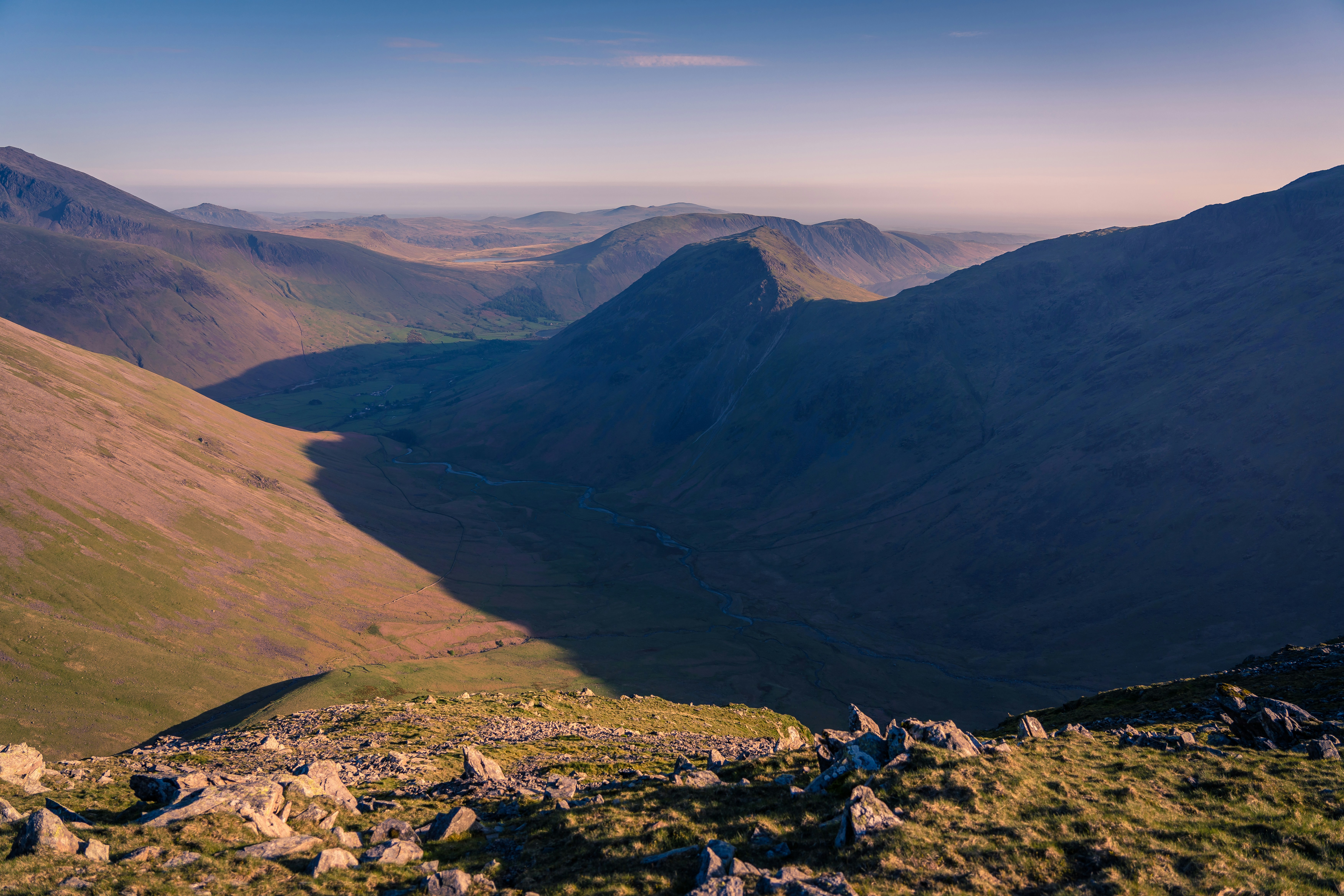 Expansive view of rolling hills and valleys under a soft sky, capturing the tranquility of nature's landscape. The interplay of light and shadow enhances the depth of the scene.