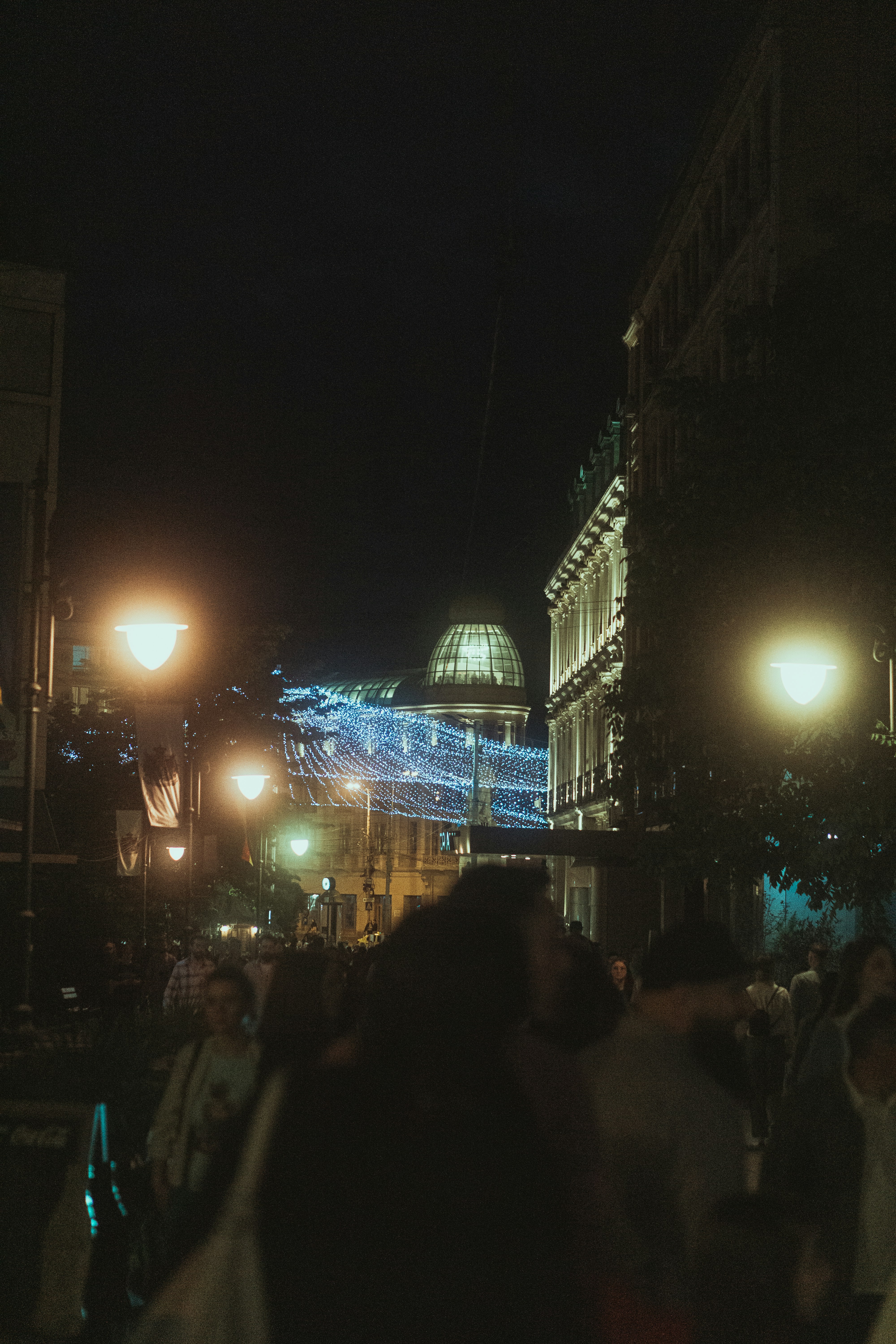 Crowd of people walking through a city street adorned with festive lights and illuminated buildings at night.