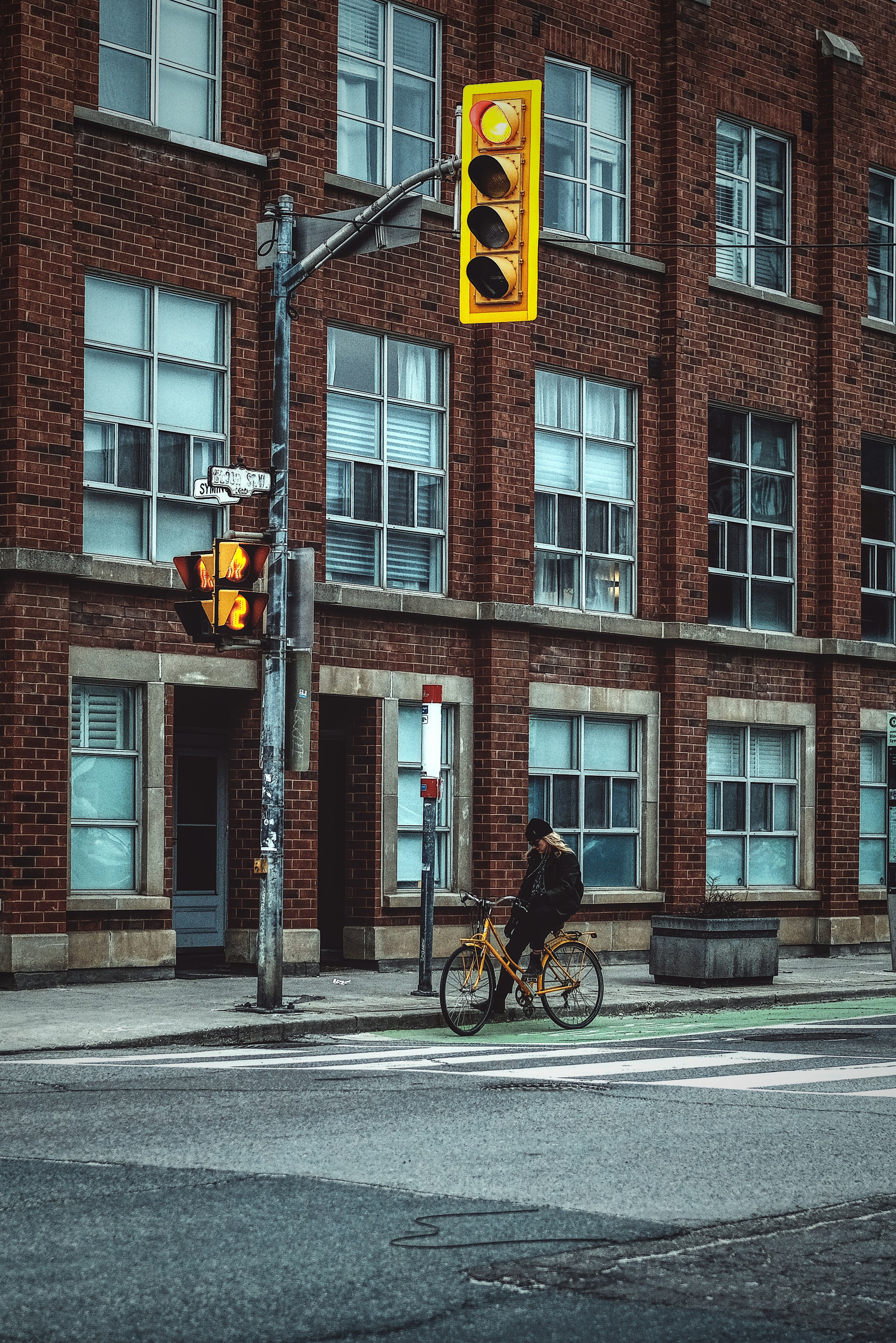 A cyclist waits at a traffic light in an urban setting, framed by red brick buildings and street signs.