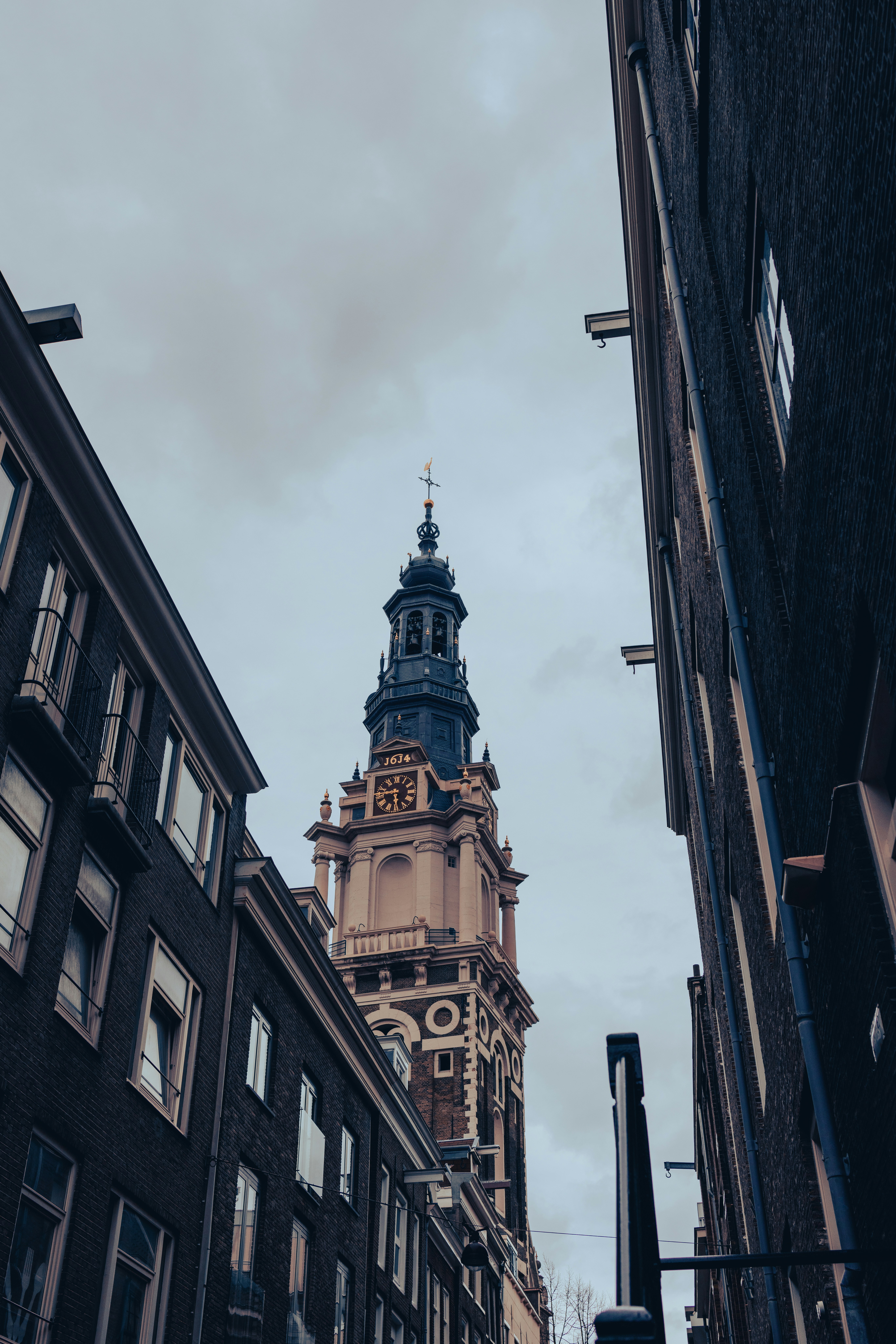 Historic clock tower rises majestically between narrow urban buildings under a cloudy sky.