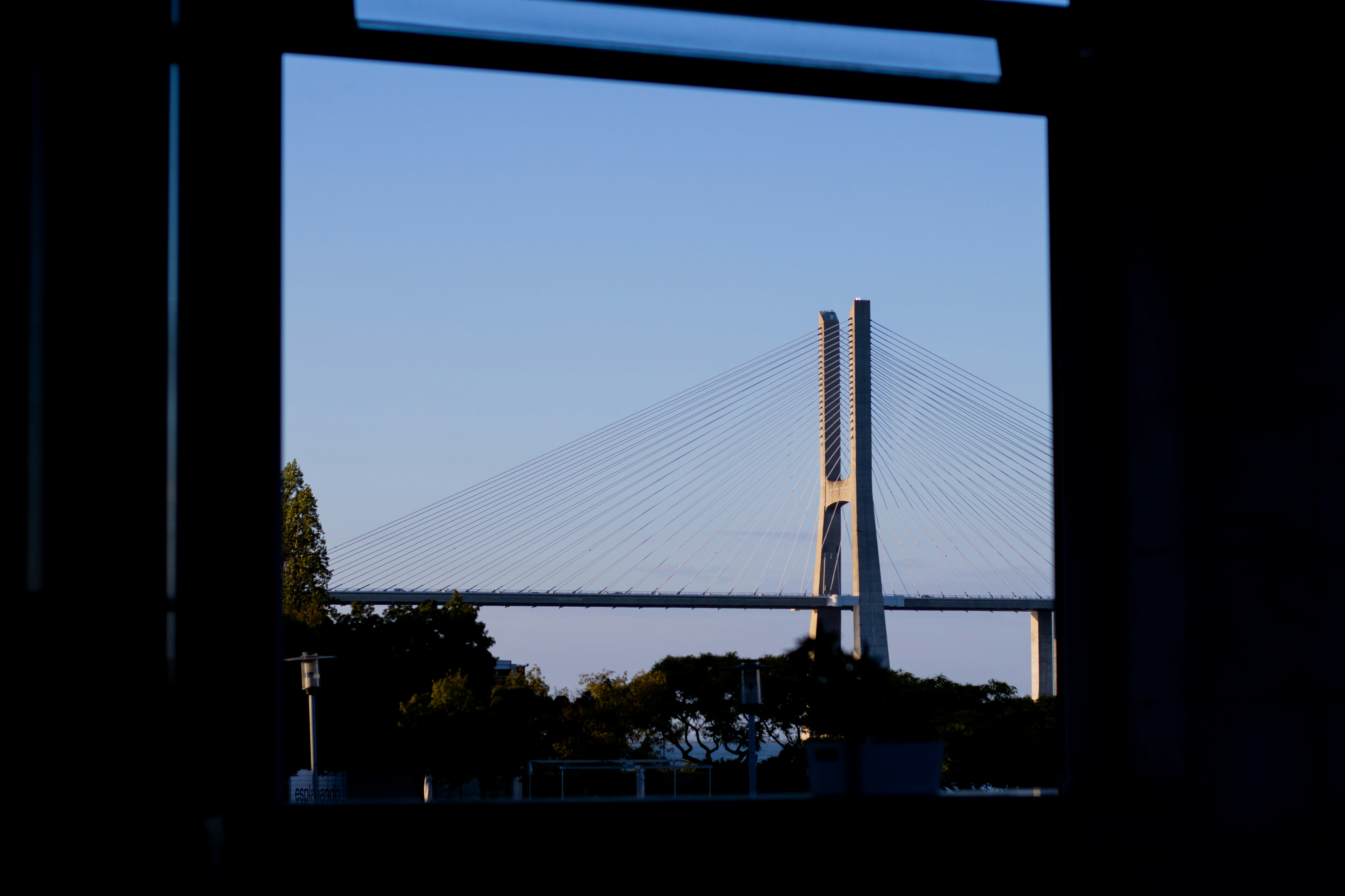 Suspension bridge framed by a window, silhouetted against a clear blue sky. The scene captures the interplay of architecture and nature.