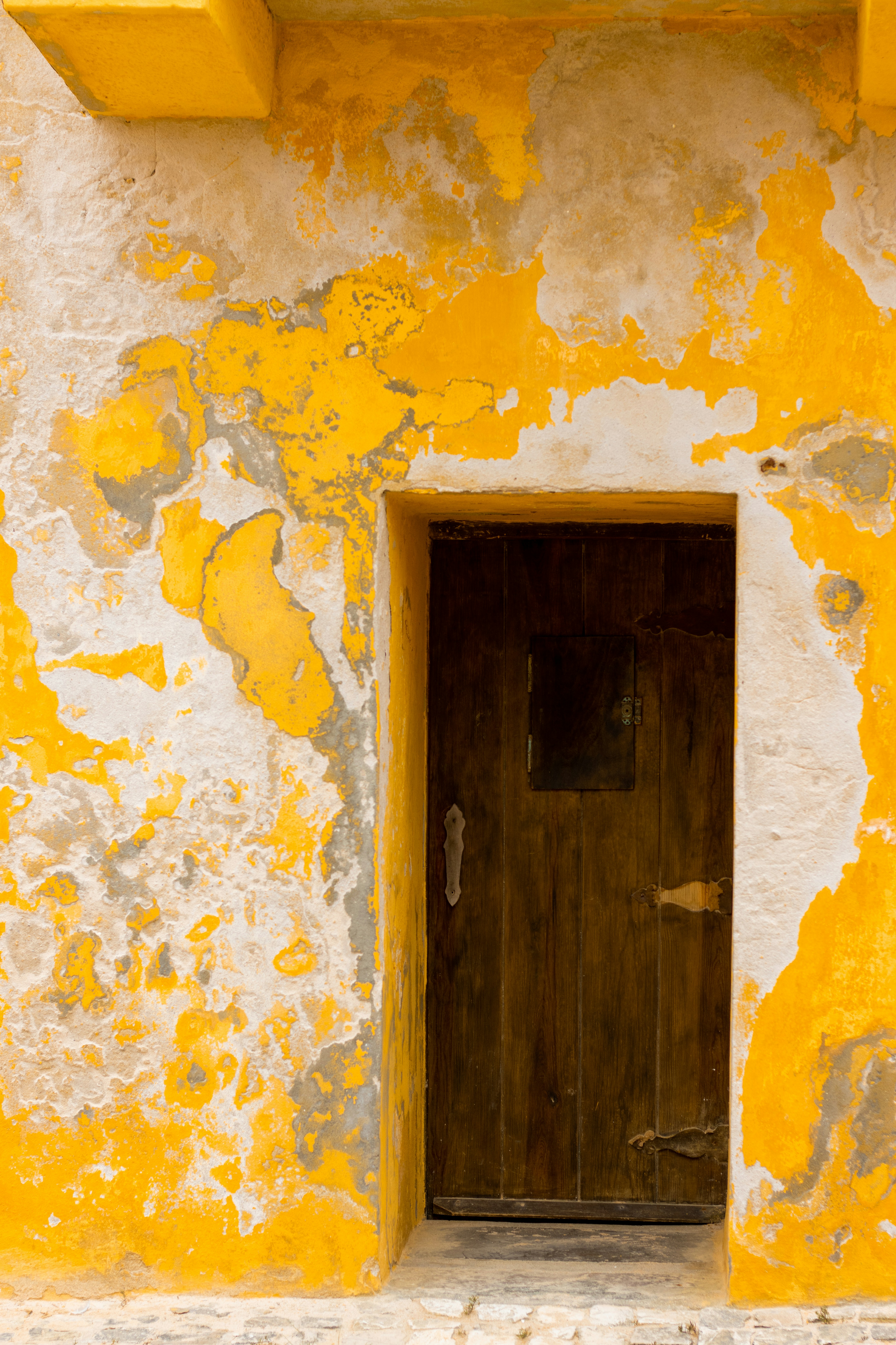 A rustic wooden door set against a vibrant yellow and peeling wall, showcasing the passage of time and texture. The door's small window adds a touch of intrigue.