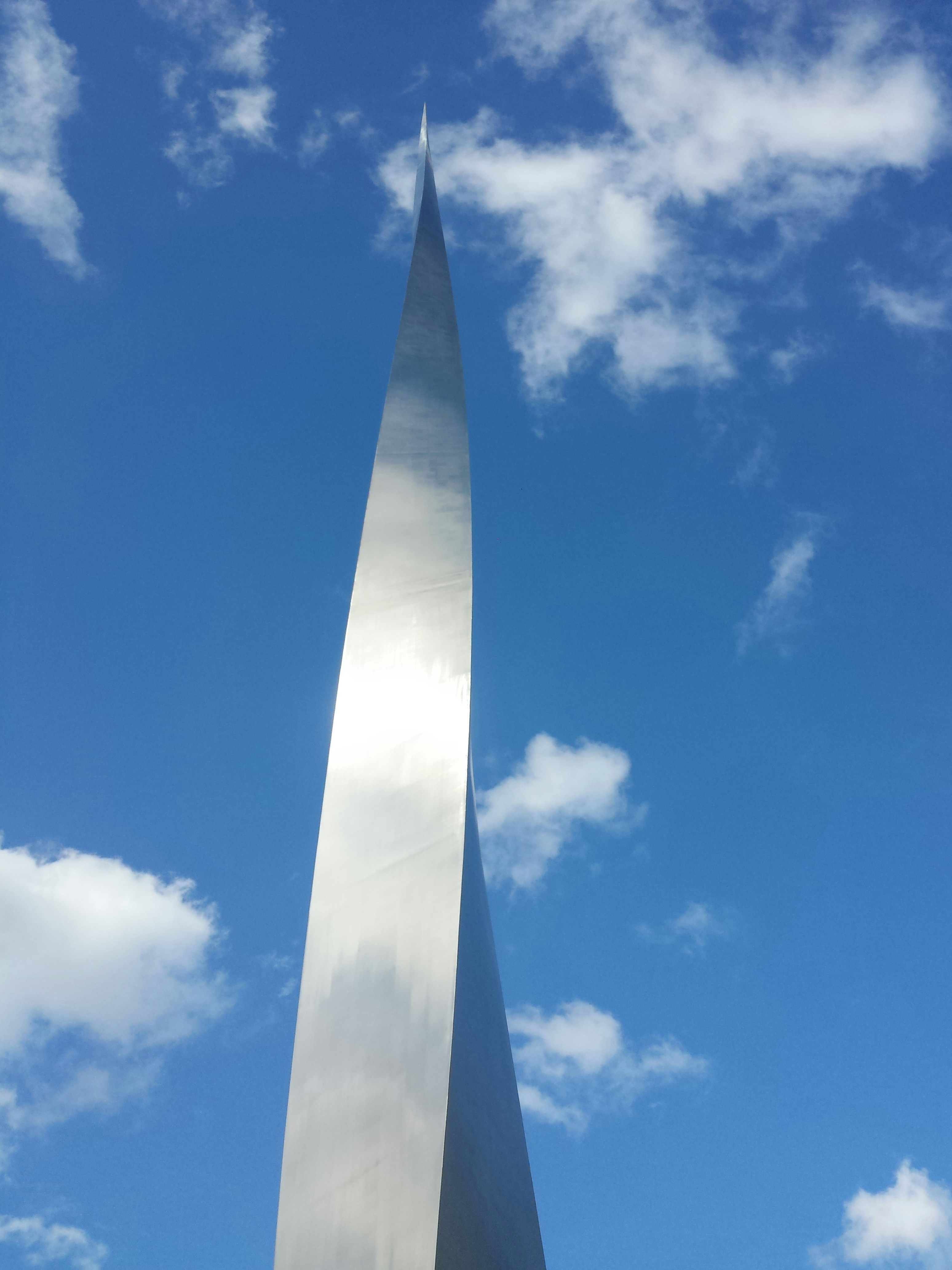 Tall stainless steel obelisk rises against a bright blue sky with scattered clouds.