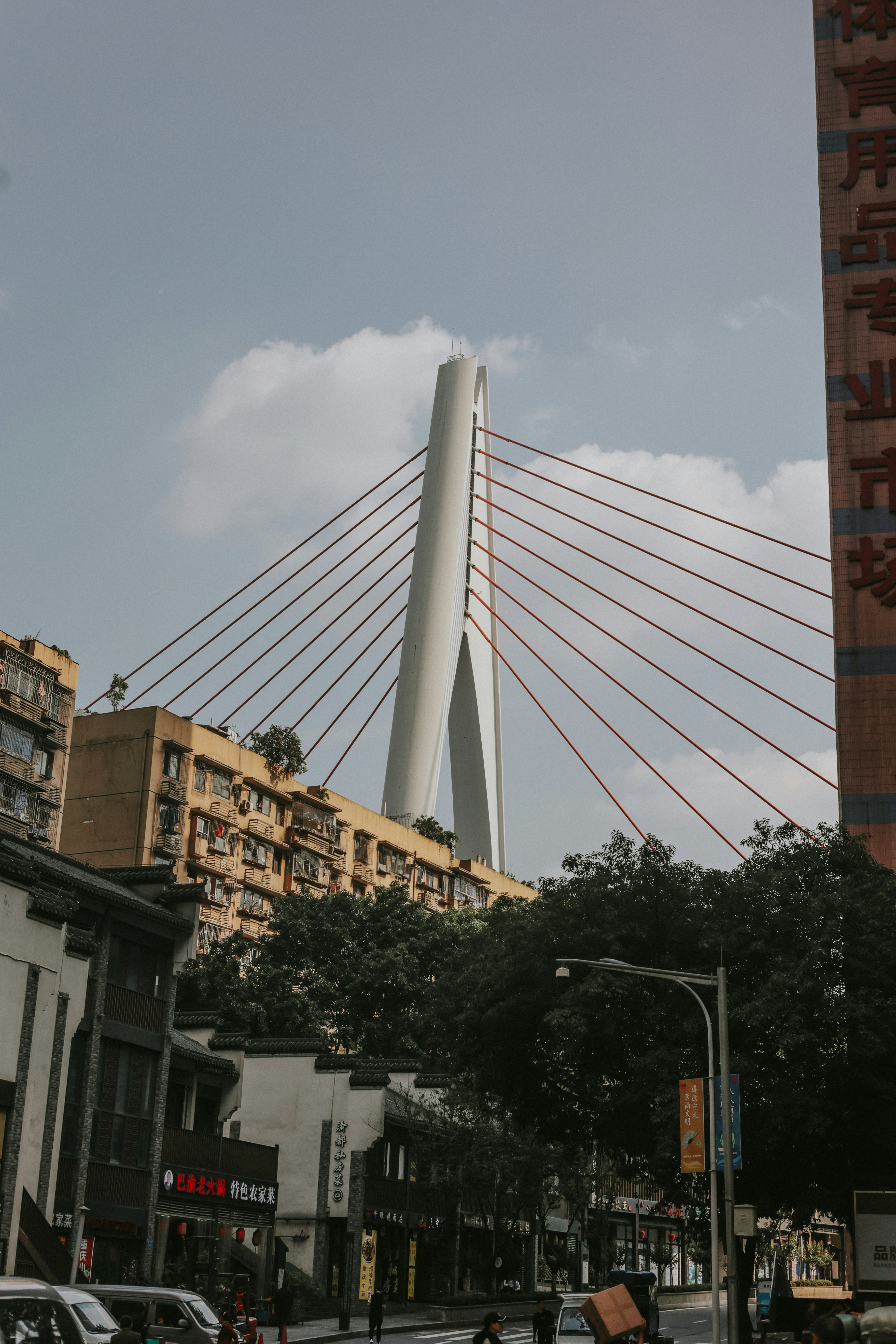 A contemporary bridge stands tall against a backdrop of urban architecture, showcasing a blend of modern engineering and historical buildings. Red cables create a striking contrast against the sky.