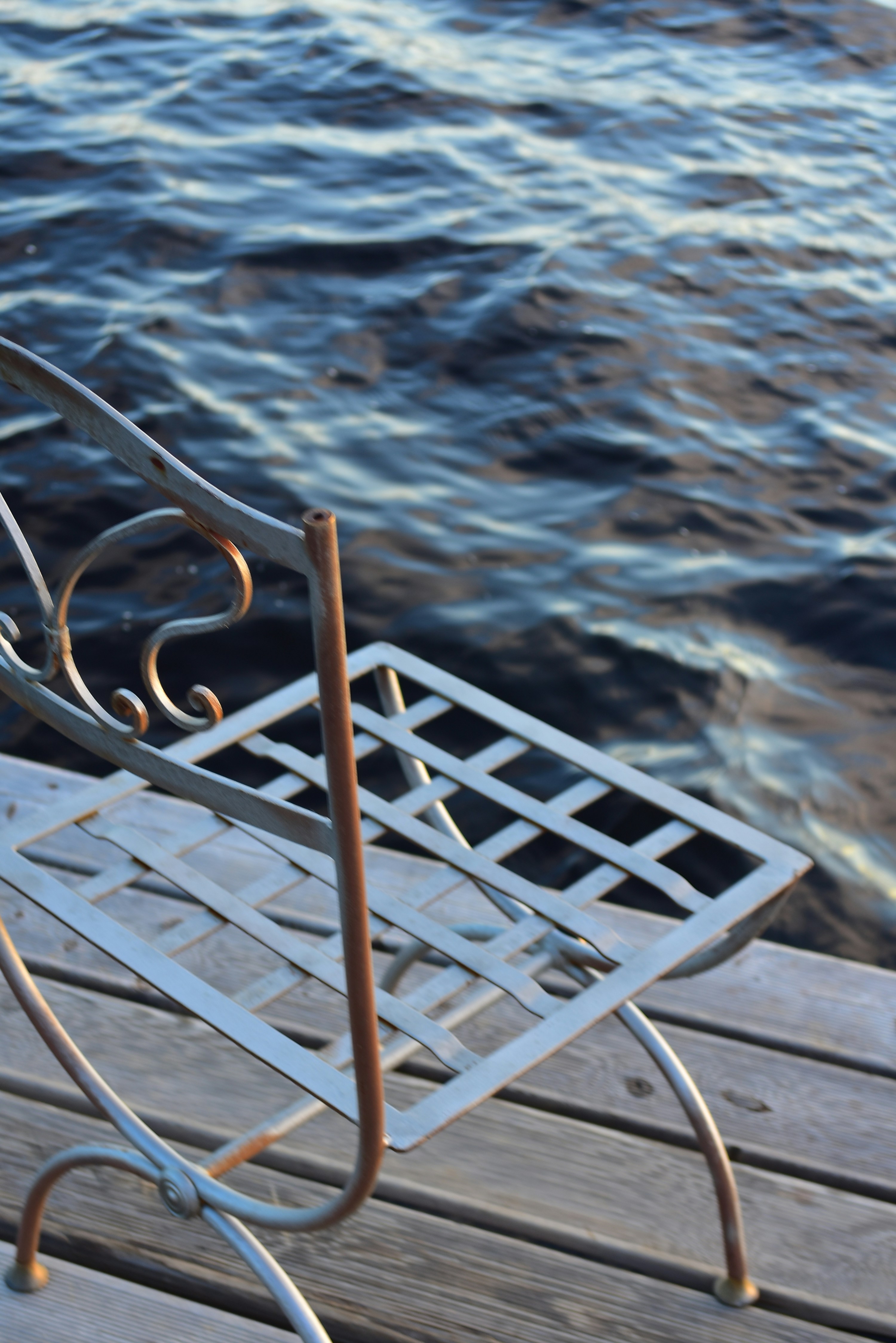 Rustic metal chair positioned on a wooden dock overlooking gently rippling water. The scene evokes a sense of tranquility and contemplation.