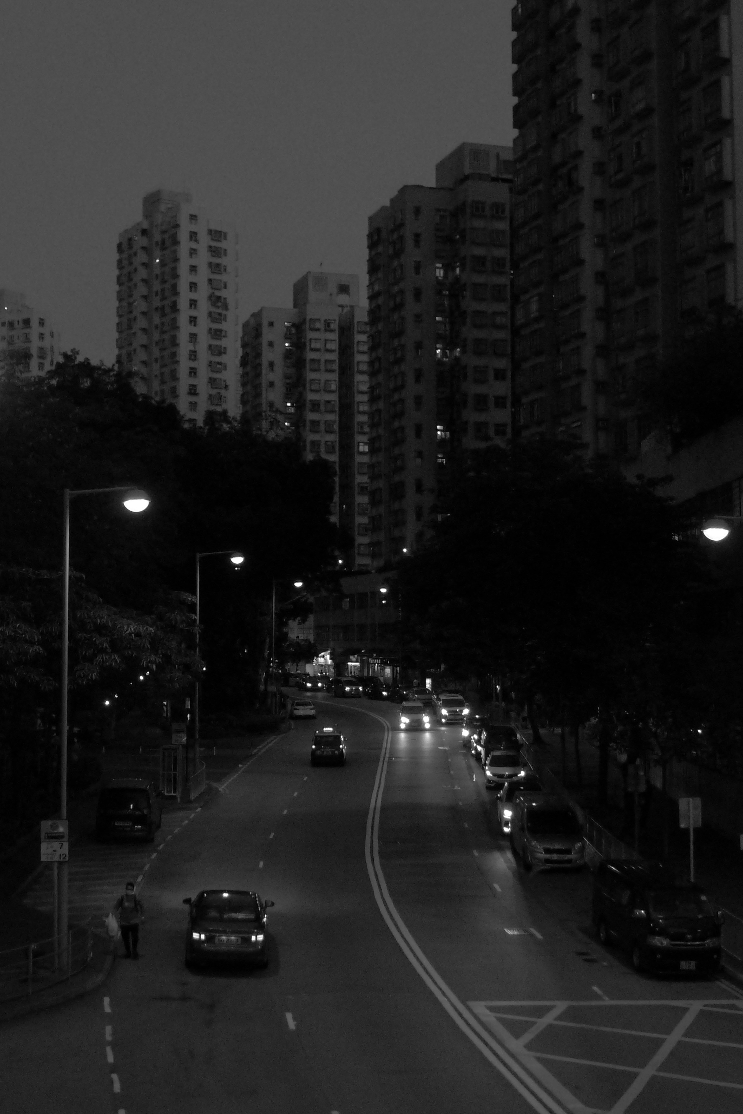 Monochrome night photograph of a curved city street lined with parked cars and towering apartment blocks, lit by streetlamps.