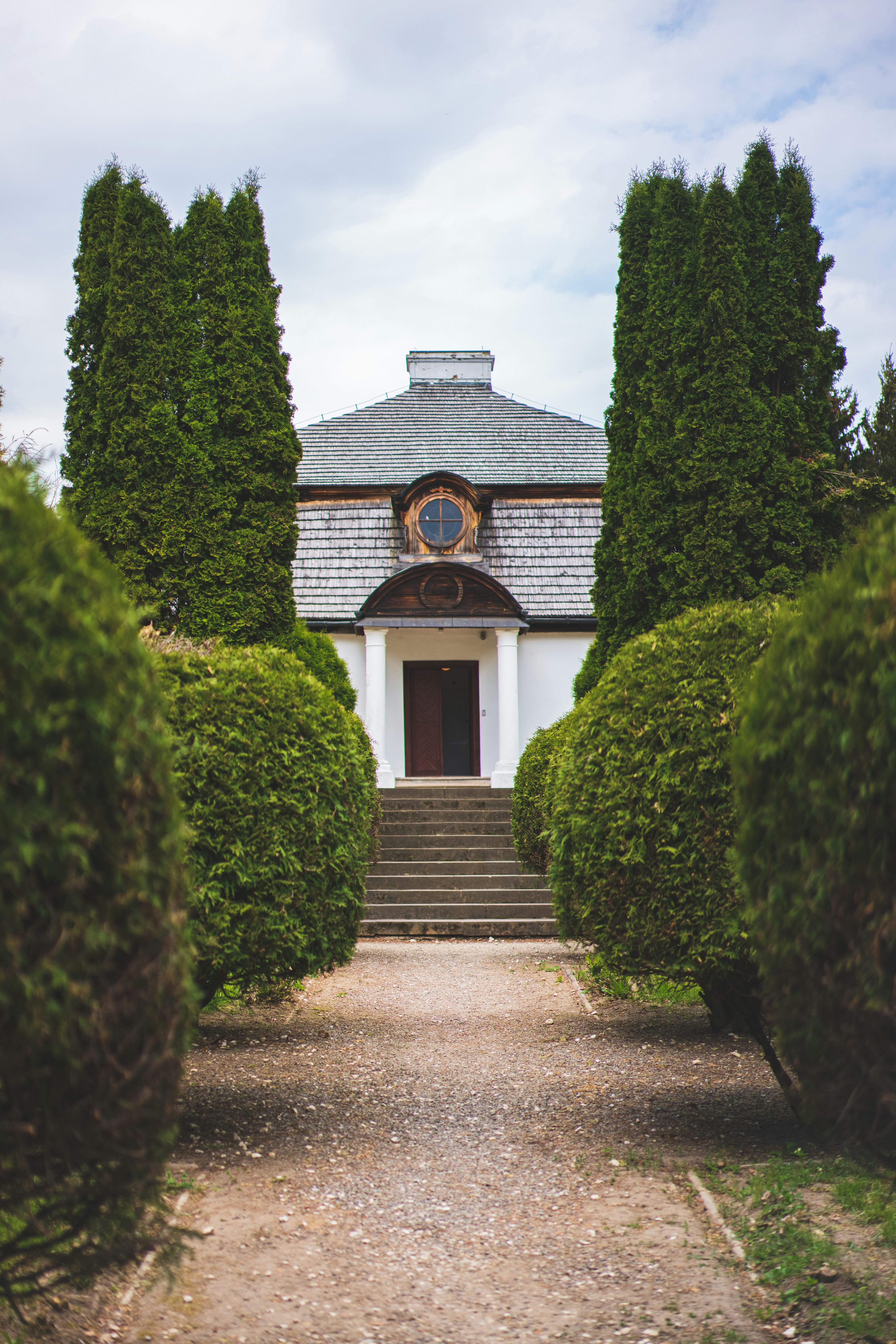 A serene pathway flanked by neatly trimmed hedges leads to a charming building with a distinctive circular window. The scene evokes tranquility and symmetry.