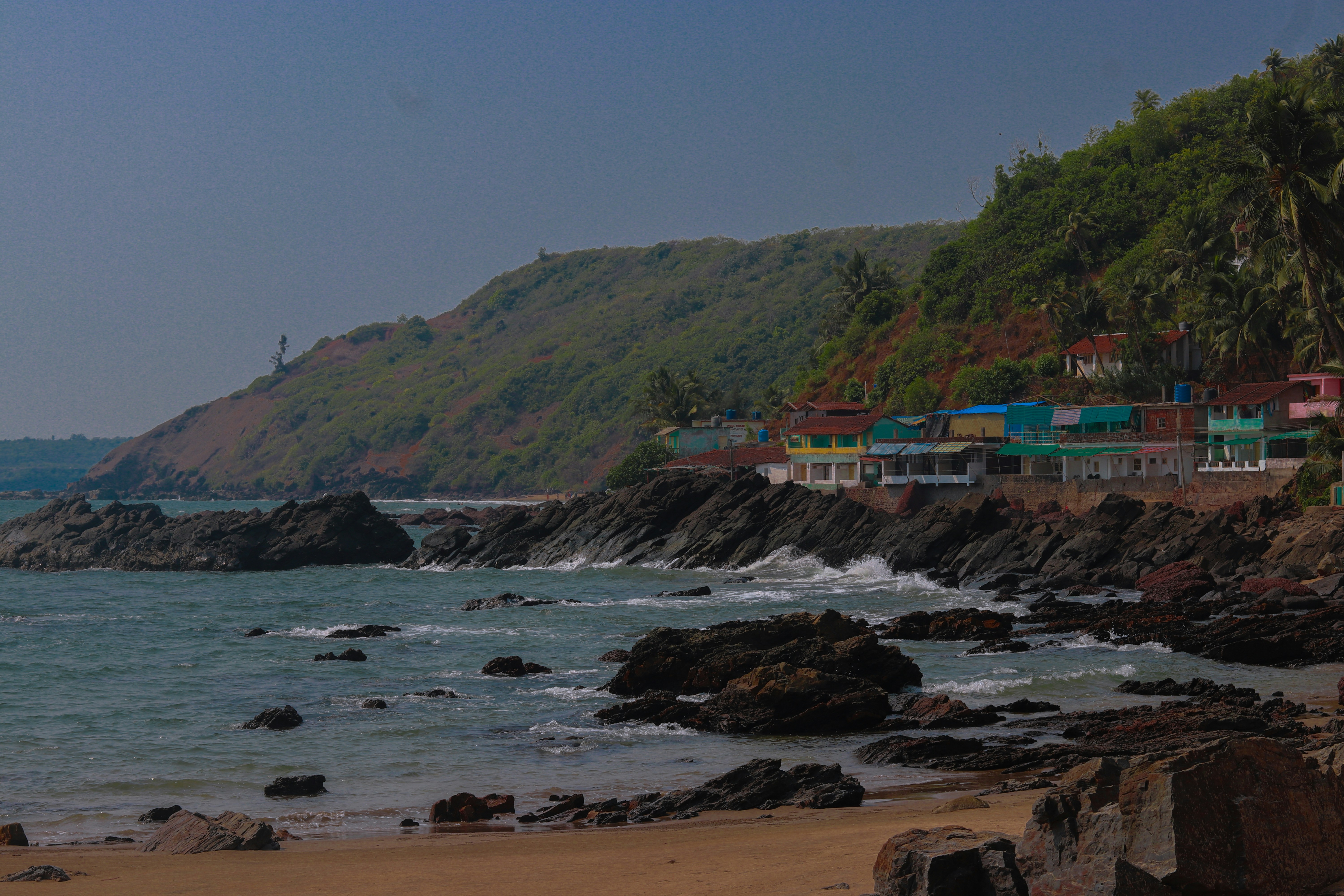 Vibrant coastal houses nestled against a lush hillside, with waves gently lapping at rocky shores. A lighthouse stands sentinel in the distance.