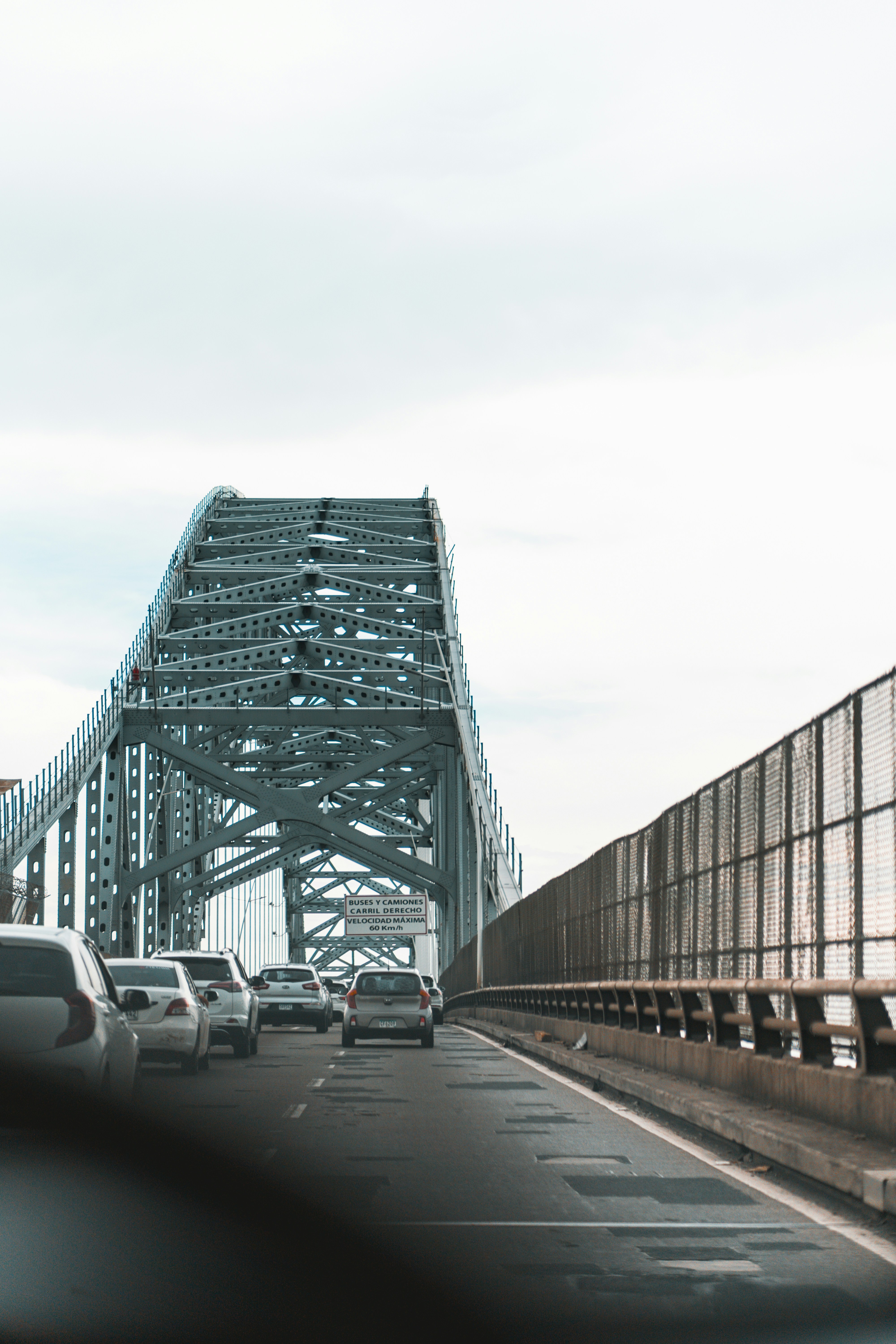 Metal bridge arching over a busy roadway, framed by cars and a protective fence. A cloudy sky looms above.