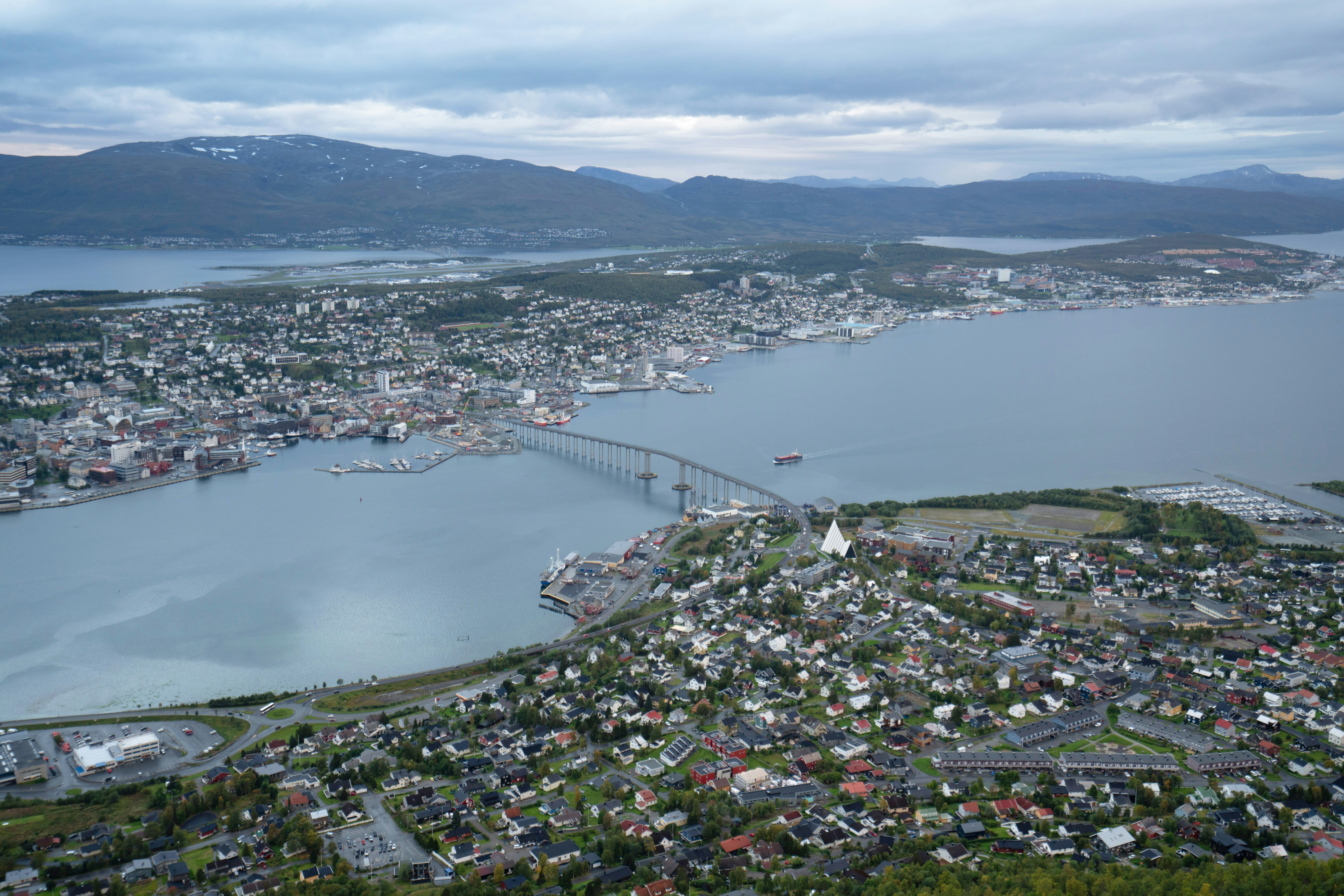 Vast aerial view of Tromsø showcasing the blend of urban architecture and natural landscapes, with a prominent bridge connecting the landmasses. The scene is framed by mountains in the background.