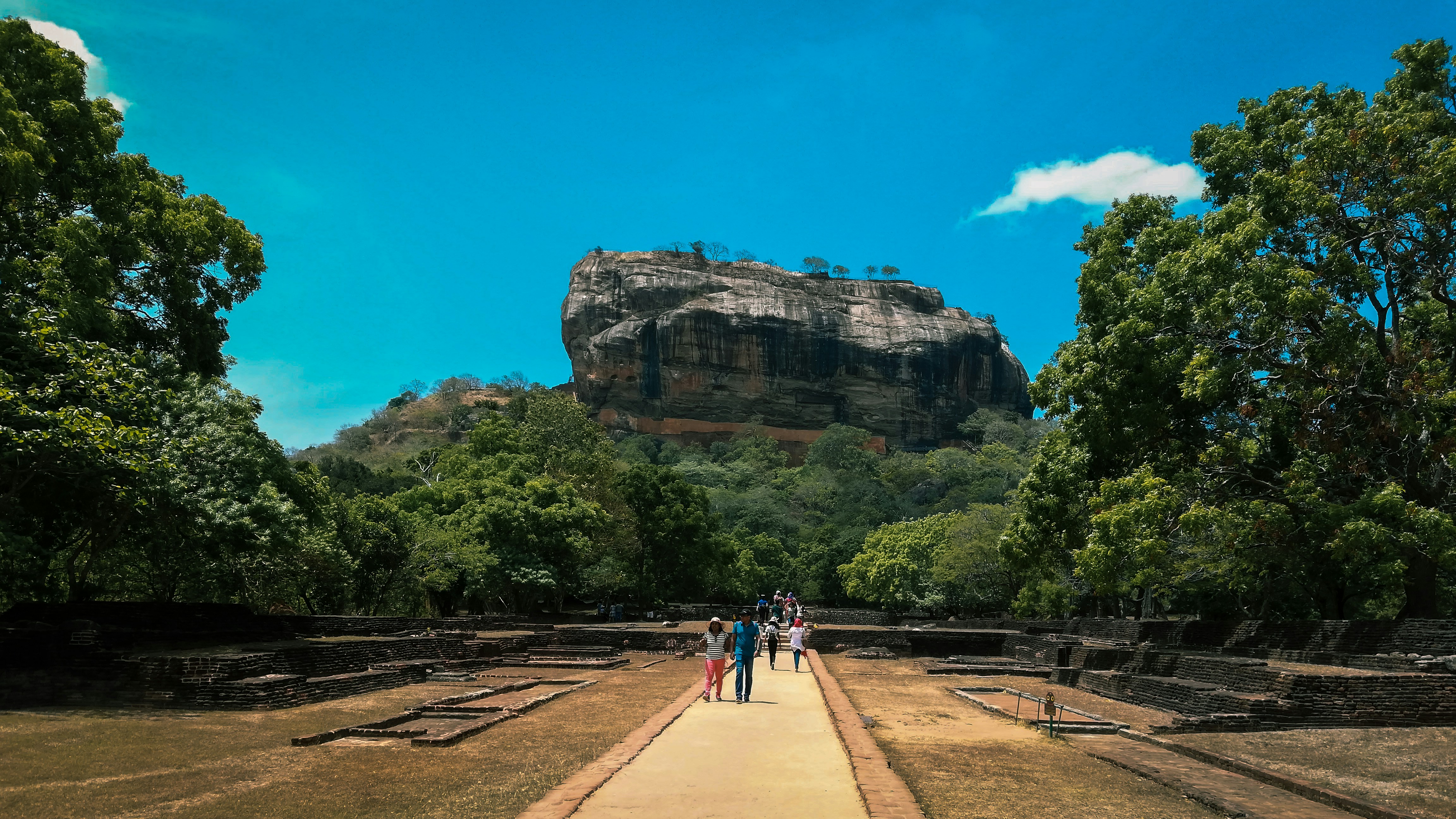 Visitors stroll along a pathway leading to a majestic rock formation, surrounded by lush greenery and ancient ruins.