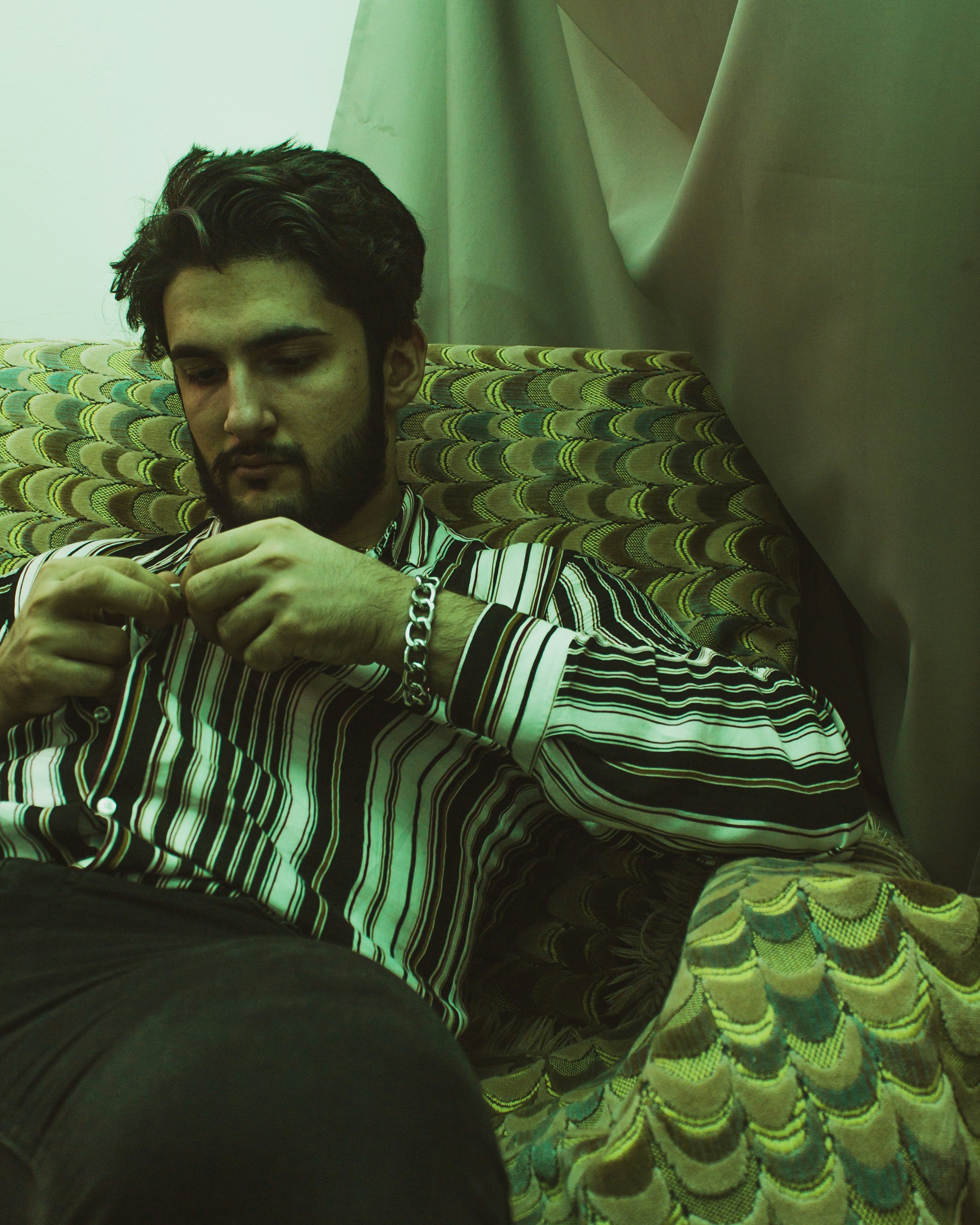 Photograph of a man in a striped shirt relaxing on a retro-patterned sofa while adjusting a bracelet. The scene is bathed in green-tinted lighting, highlighting a contemplative mood.