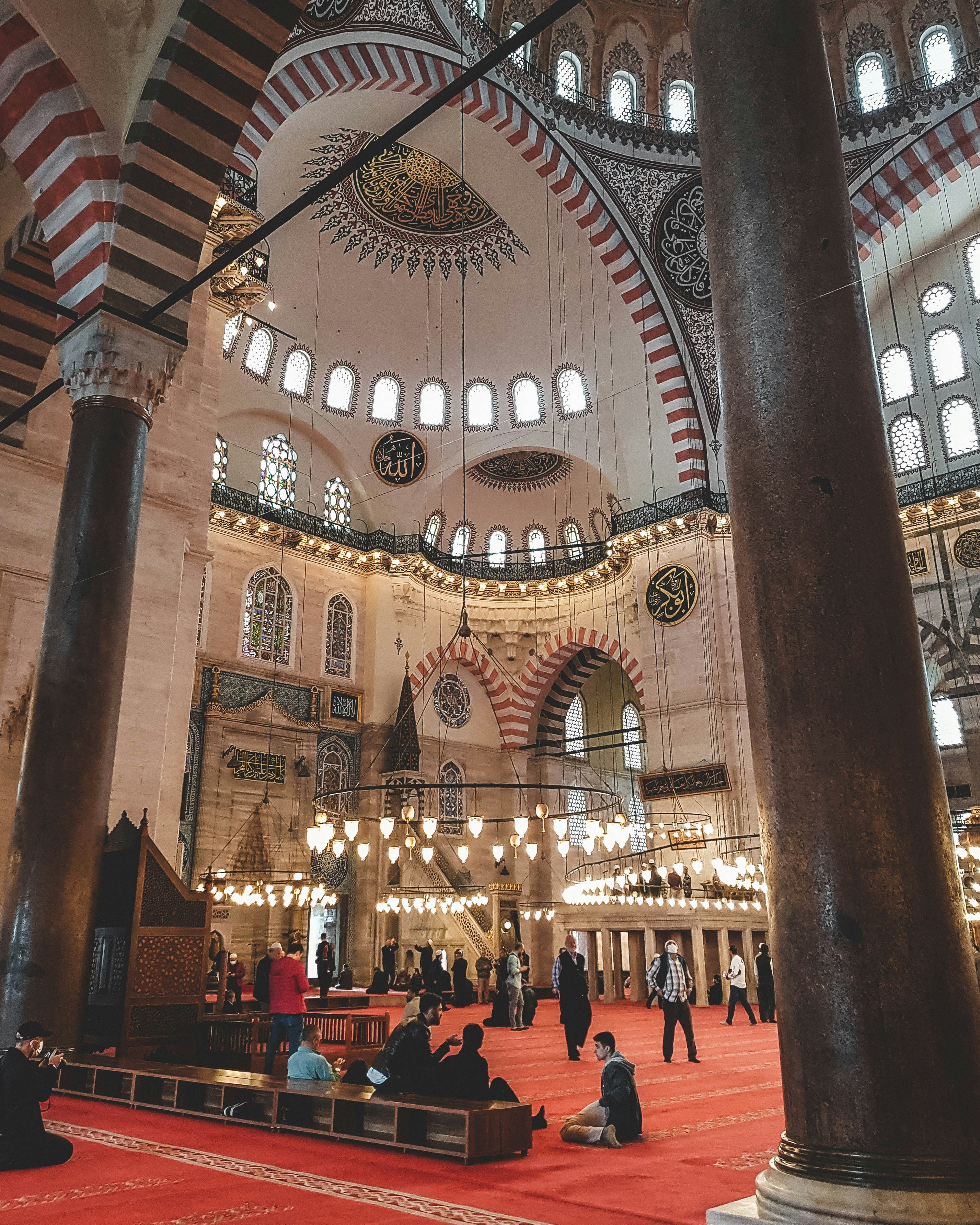 Interior view of a grand mosque showcasing its intricate domes, arches, and decorative elements, with visitors engaged in quiet reflection. 