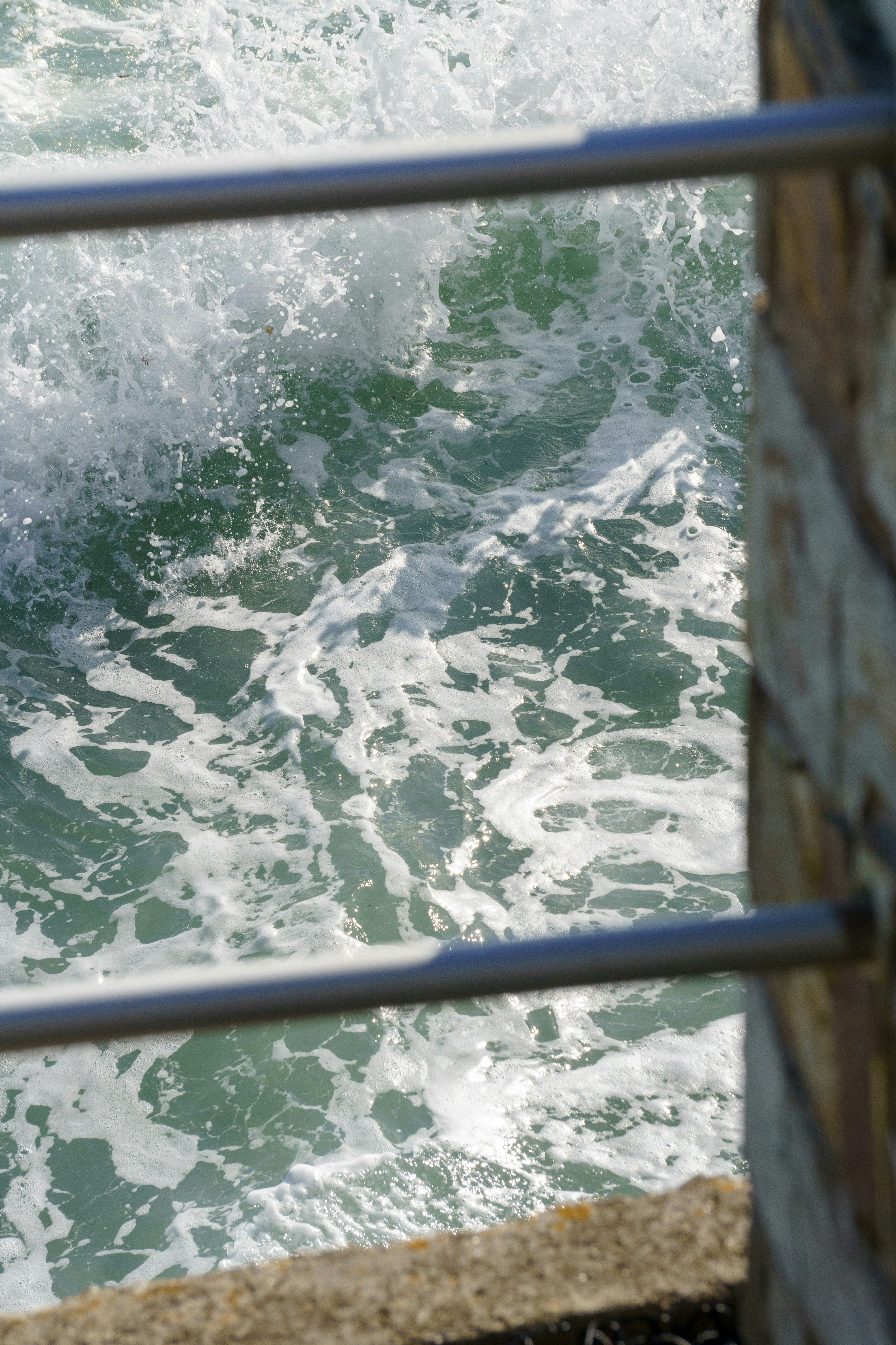 Foamy ocean waves crashing against a rocky shoreline, partially framed by a metal railing. The interplay of light and water creates a dynamic scene.