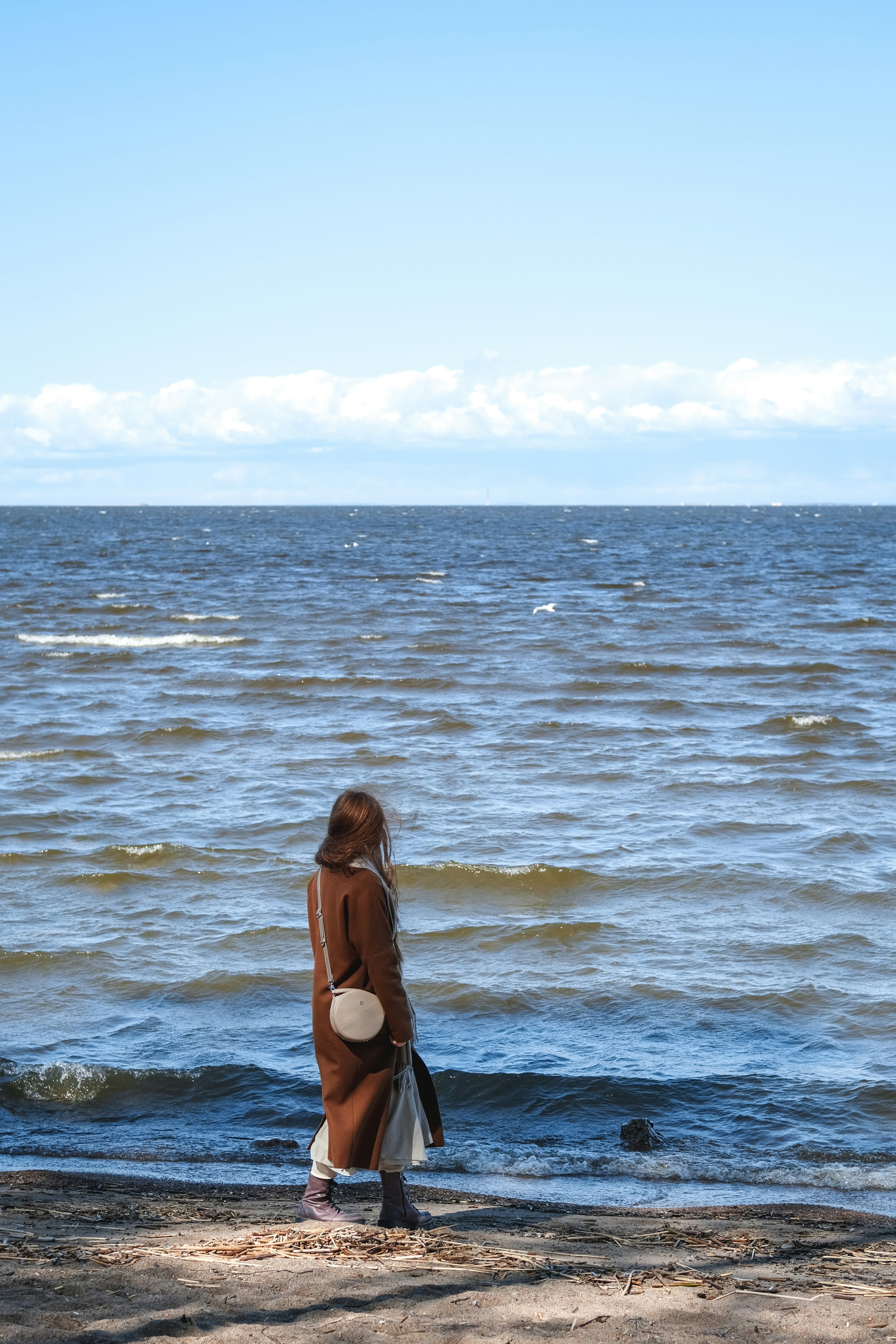 A woman stands thoughtfully at the shoreline, gazing out over rippling waves under a clear blue sky.