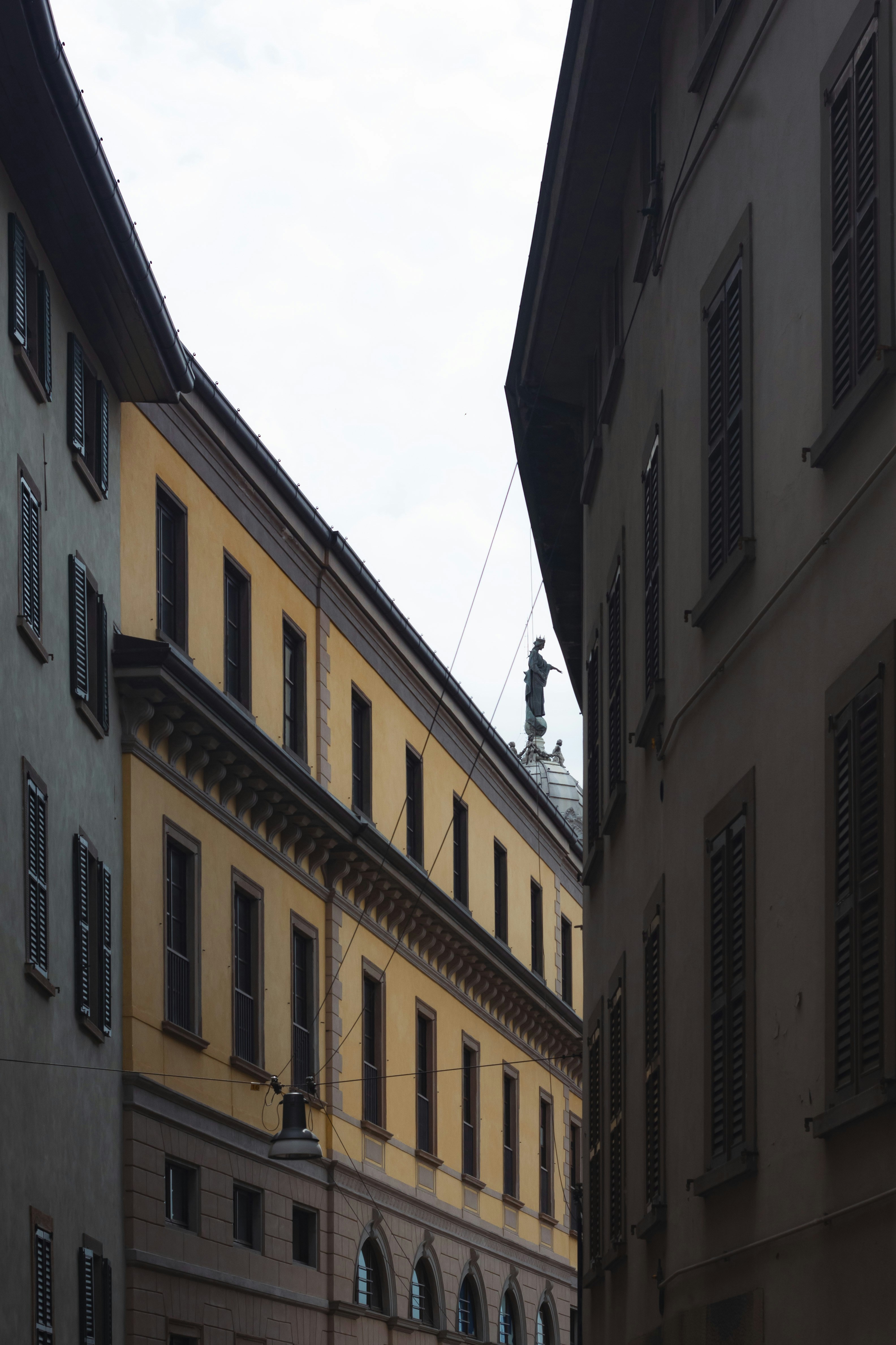 Narrow alley framed by historic buildings, revealing a statue atop a distant structure. The interplay of light and architecture creates a sense of depth.