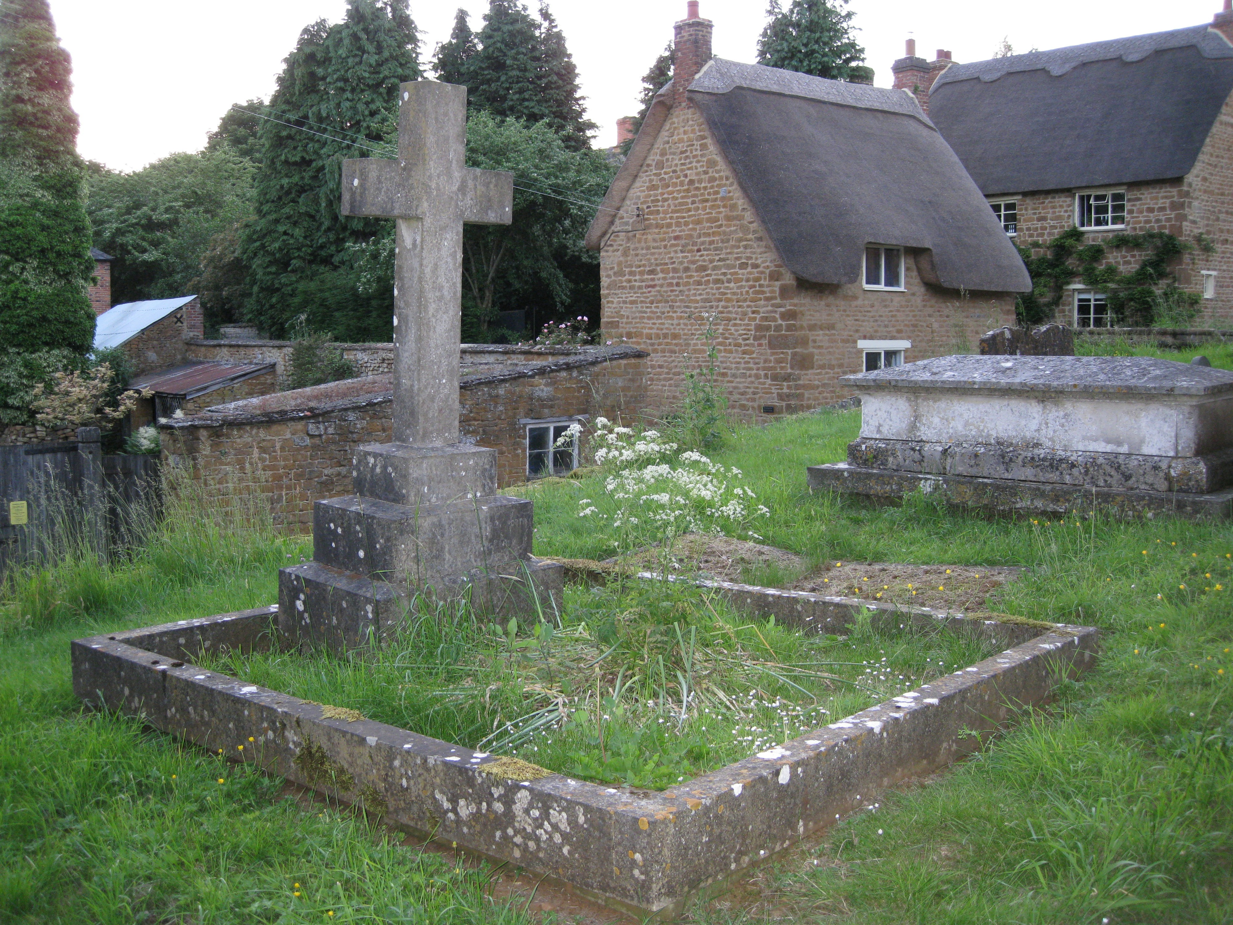 Weathered gravestone surrounded by overgrown grass and wildflowers, set against a backdrop of rustic cottages.