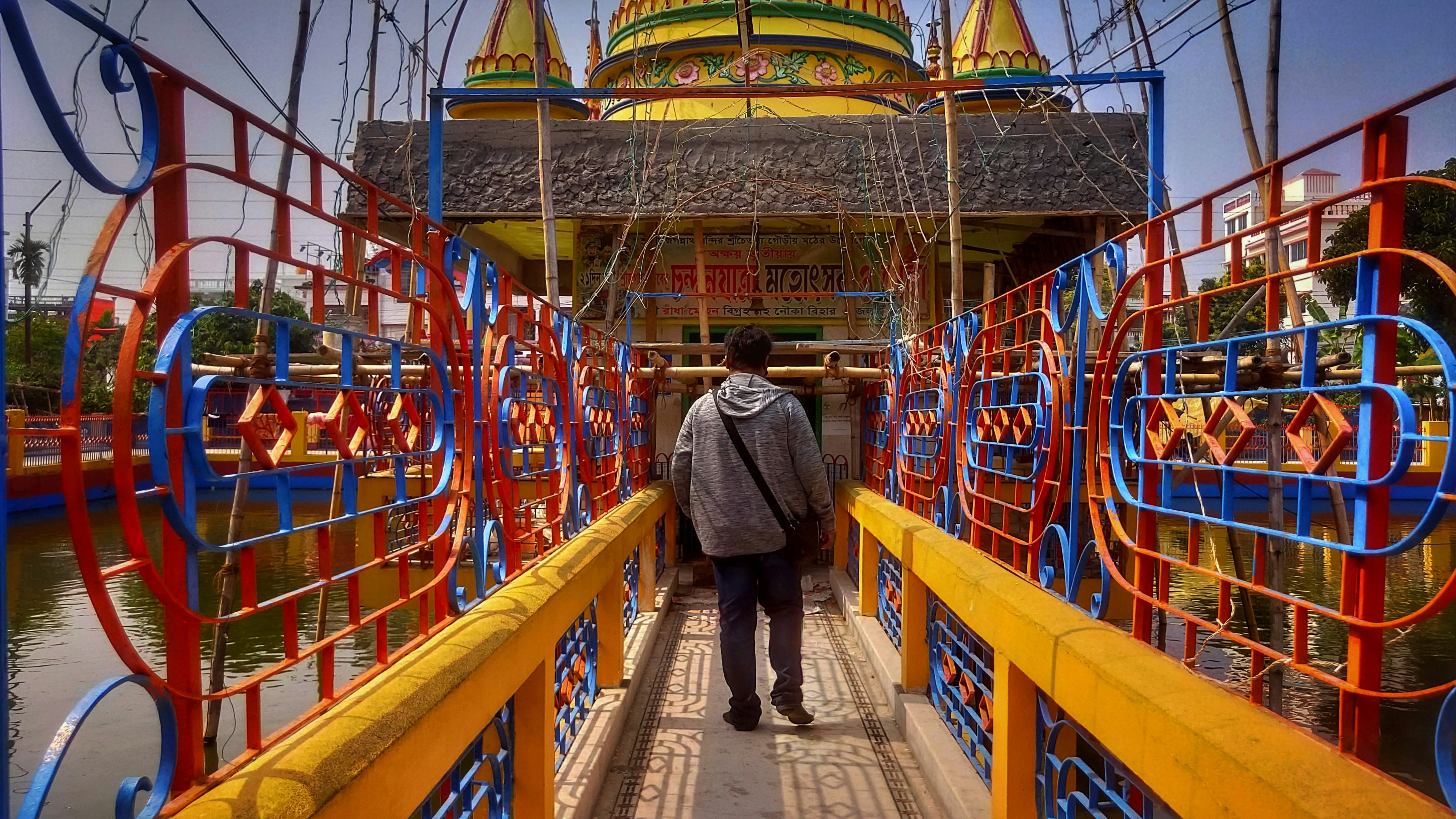 A person walks along a vibrant bridge adorned with intricate patterns, leading to a colorful temple. The scene captures a blend of culture and architecture.