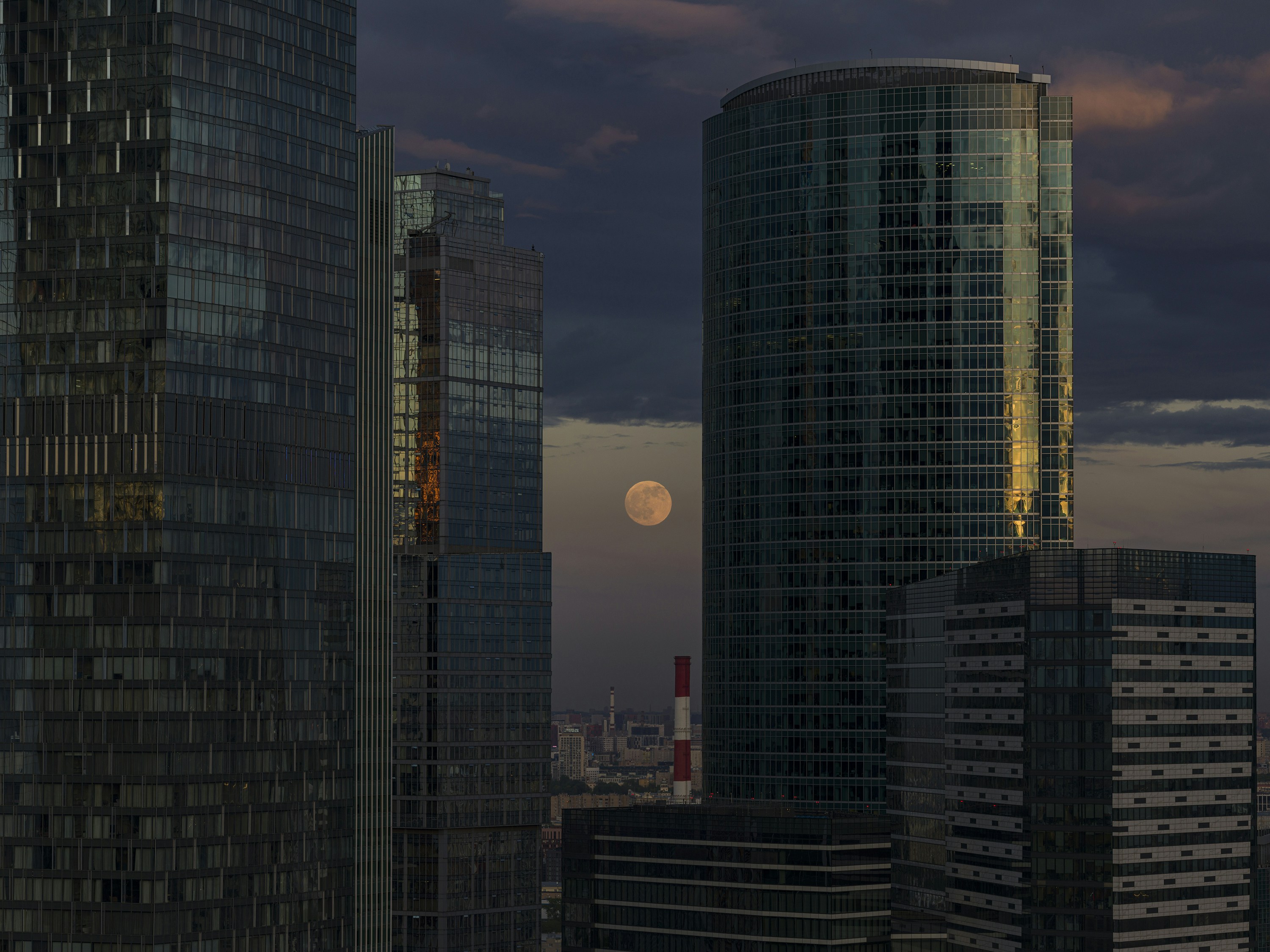 Night cityscape photograph showing the moon between tall glass towers, framed by the dense skyline. It highlights vertical geometry and the warm, dusky tones of the scene.