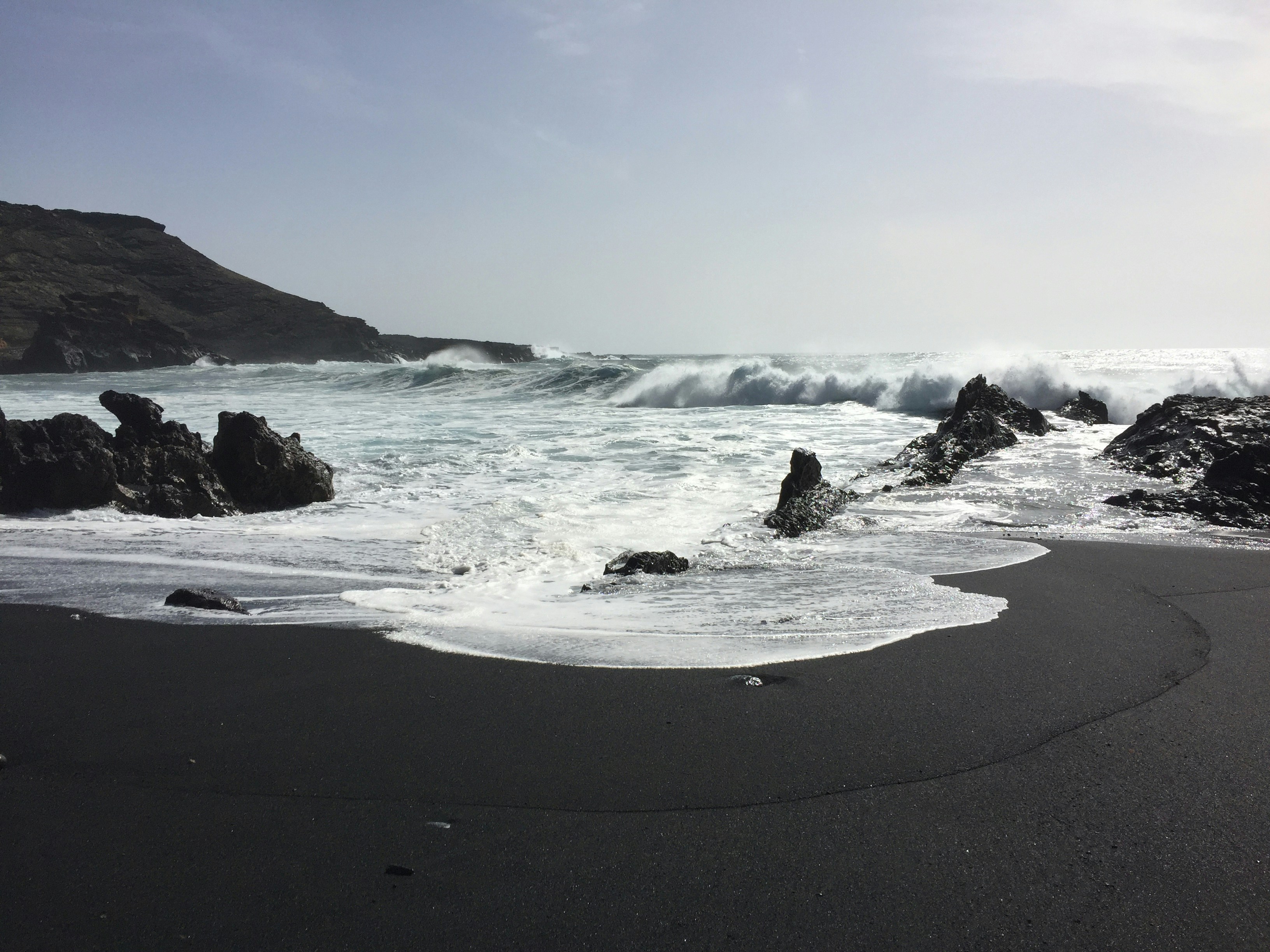 Waves crash onto a black sand beach, framed by rugged rocks and a distant hillside under a clear sky.