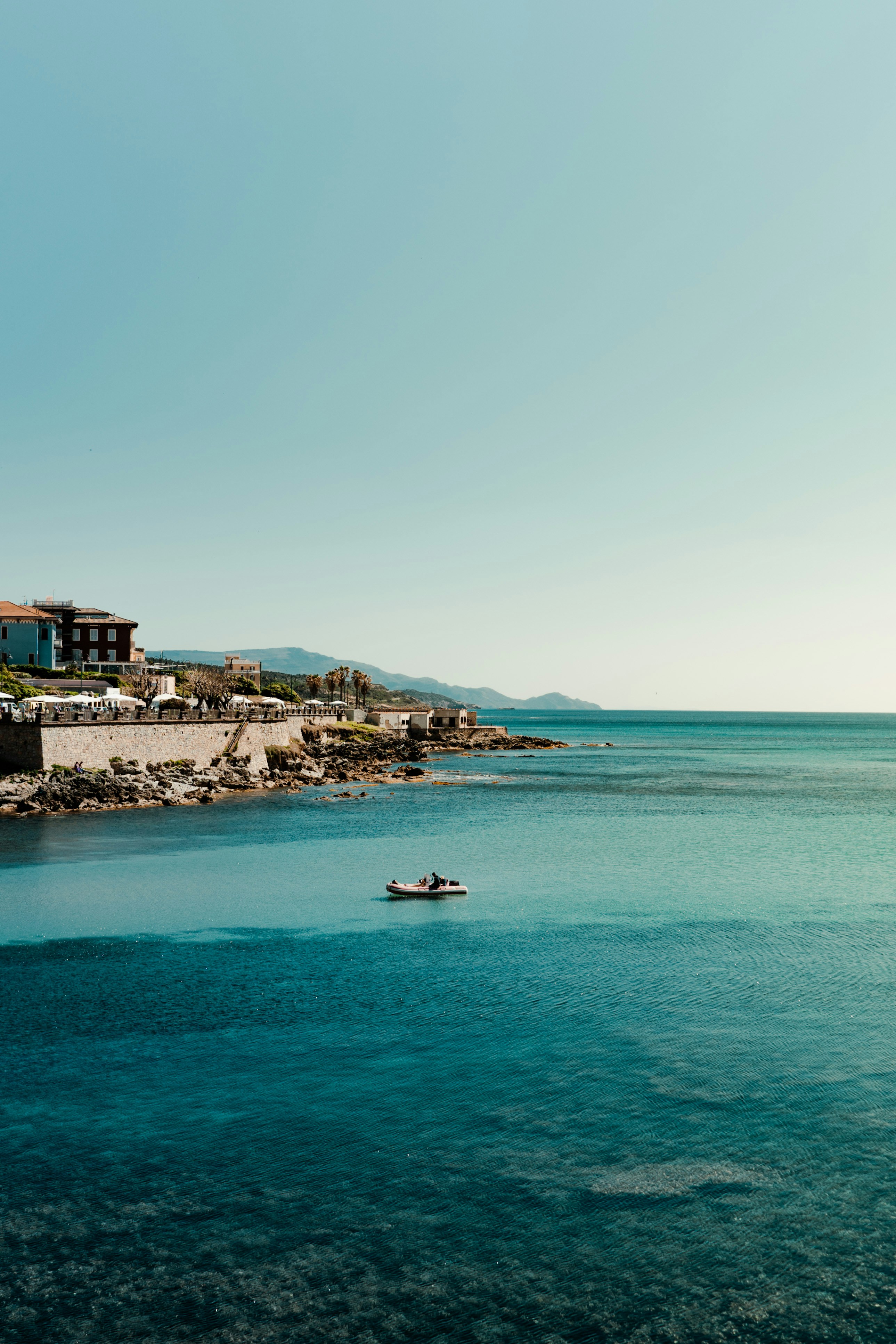 A small boat glides across tranquil turquoise waters, framed by a coastal town and distant mountains under a clear sky.