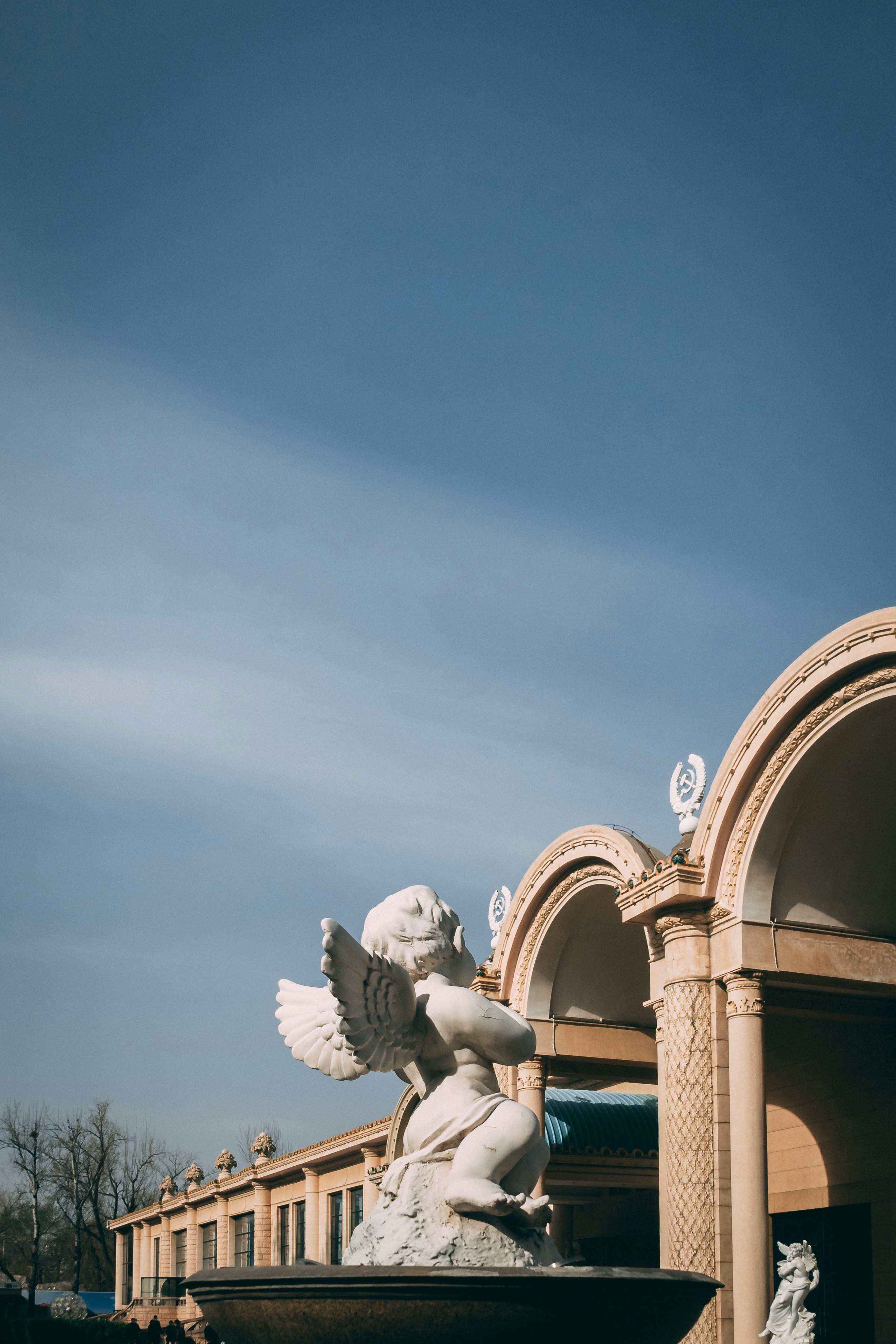 Cherubic statue with wings perched atop a fountain, framed by classical architecture under a clear blue sky.
