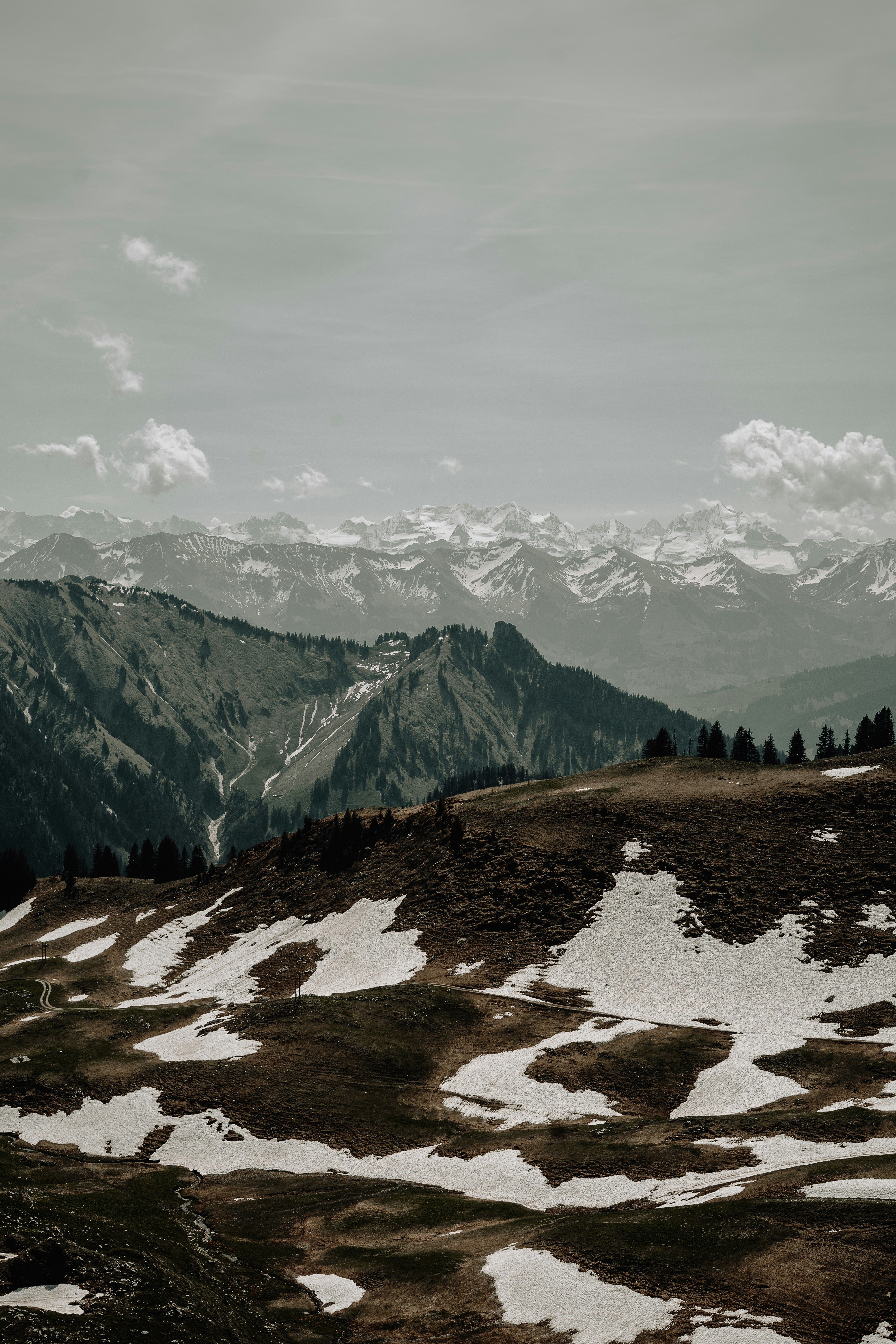 Rolling hills dotted with patches of snow and framed by majestic mountain ranges under a cloudy sky.