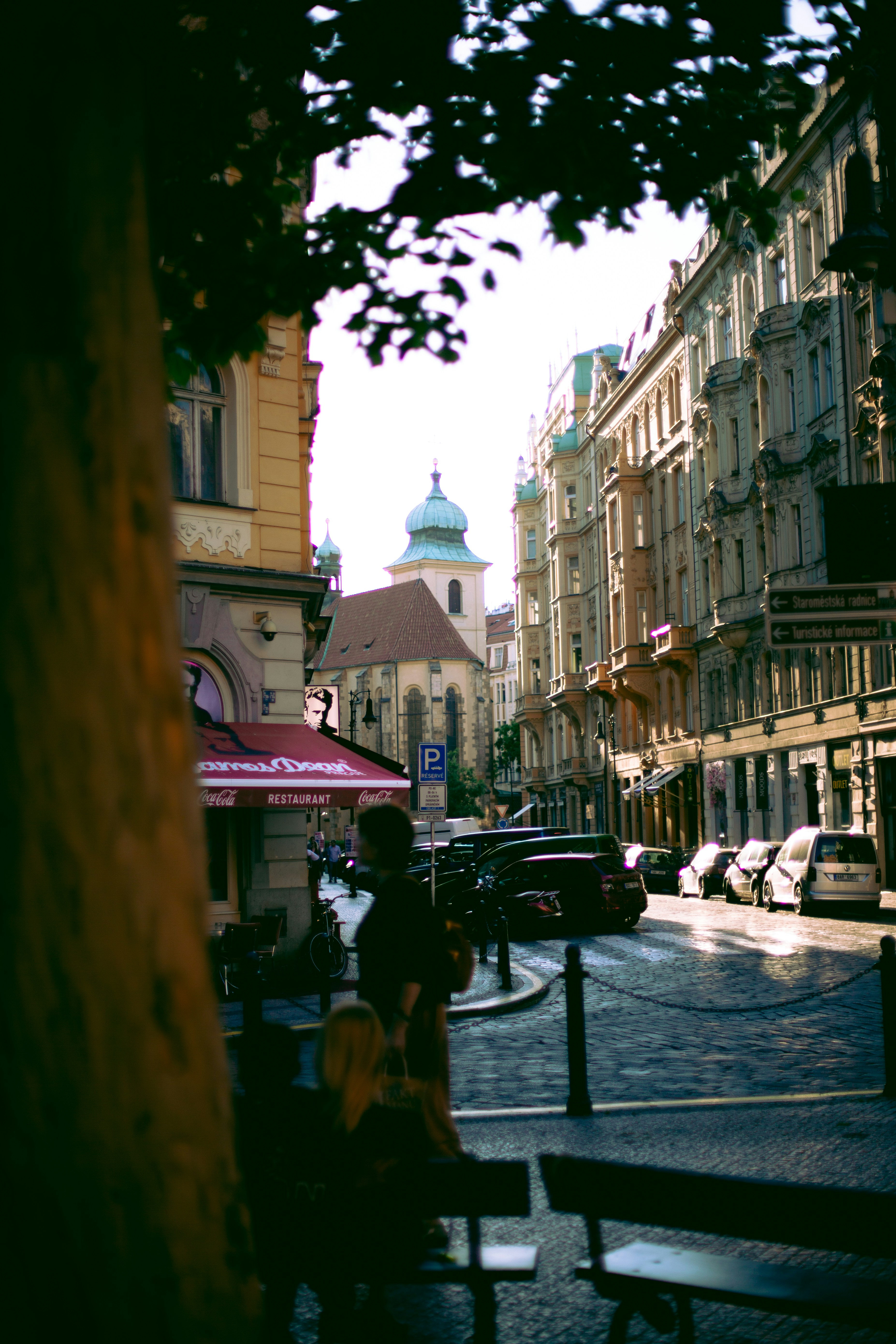 Charming cobblestone street lined with historical buildings, featuring a glimpse of a church dome in the background. A couple of figures add life to the scene.