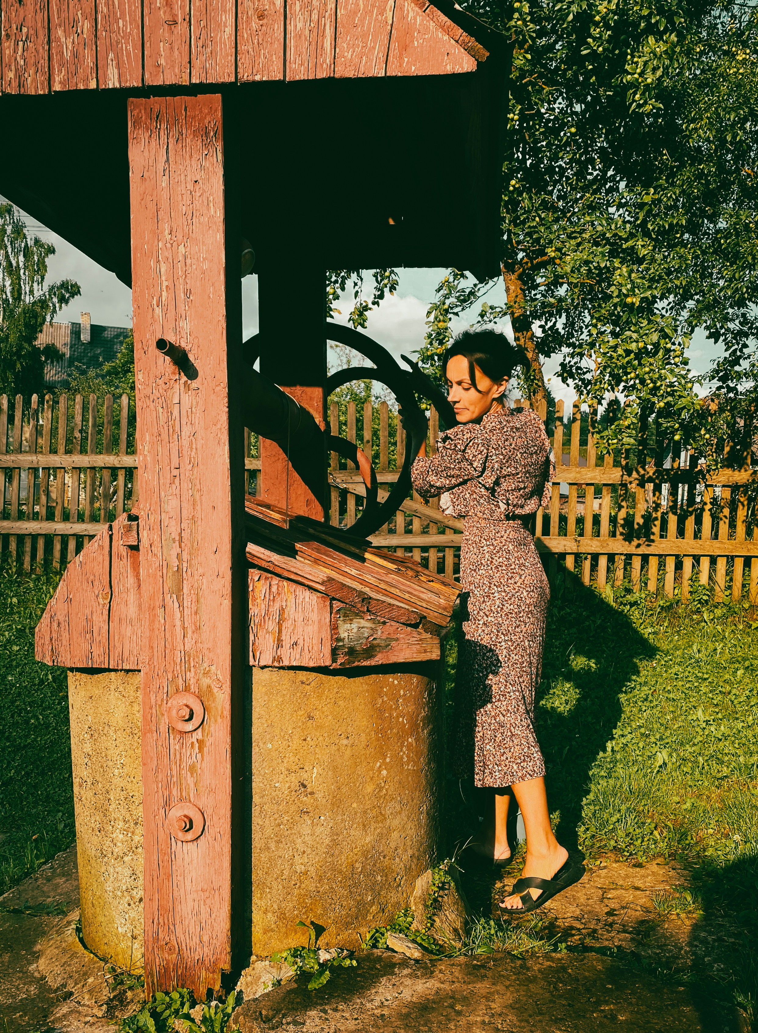 Sunlit Portrait by the Rustic Well: A Floral-Dress Woman at the Old Wooden Wheel