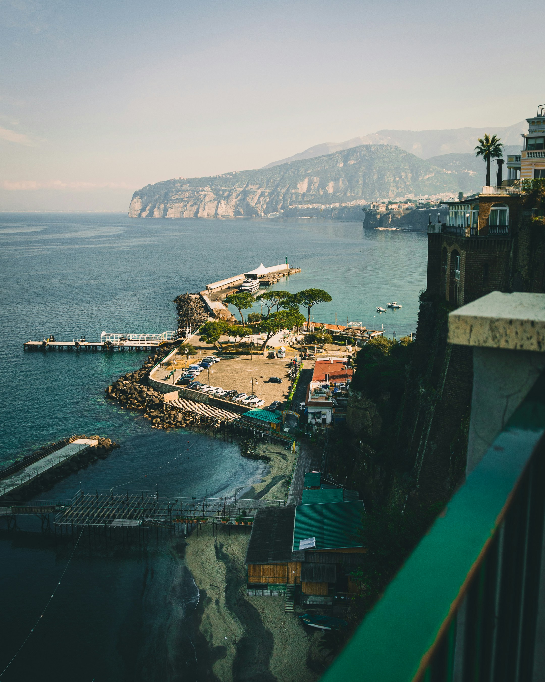 A picturesque harbor scene showcasing boats docked along the coastline, framed by lush greenery and rocky cliffs. The tranquil waters reflect the soft hues of the sky.