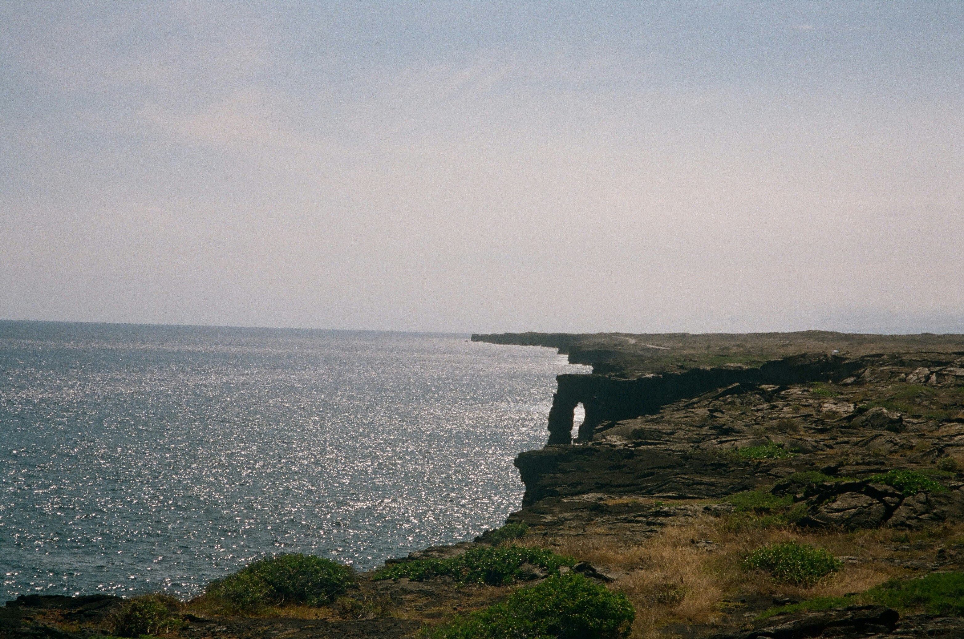 Coastal cliffs frame a natural rock arch overlooking a calm, sunlit sea. The scene emphasizes rugged geology and a broad horizon.