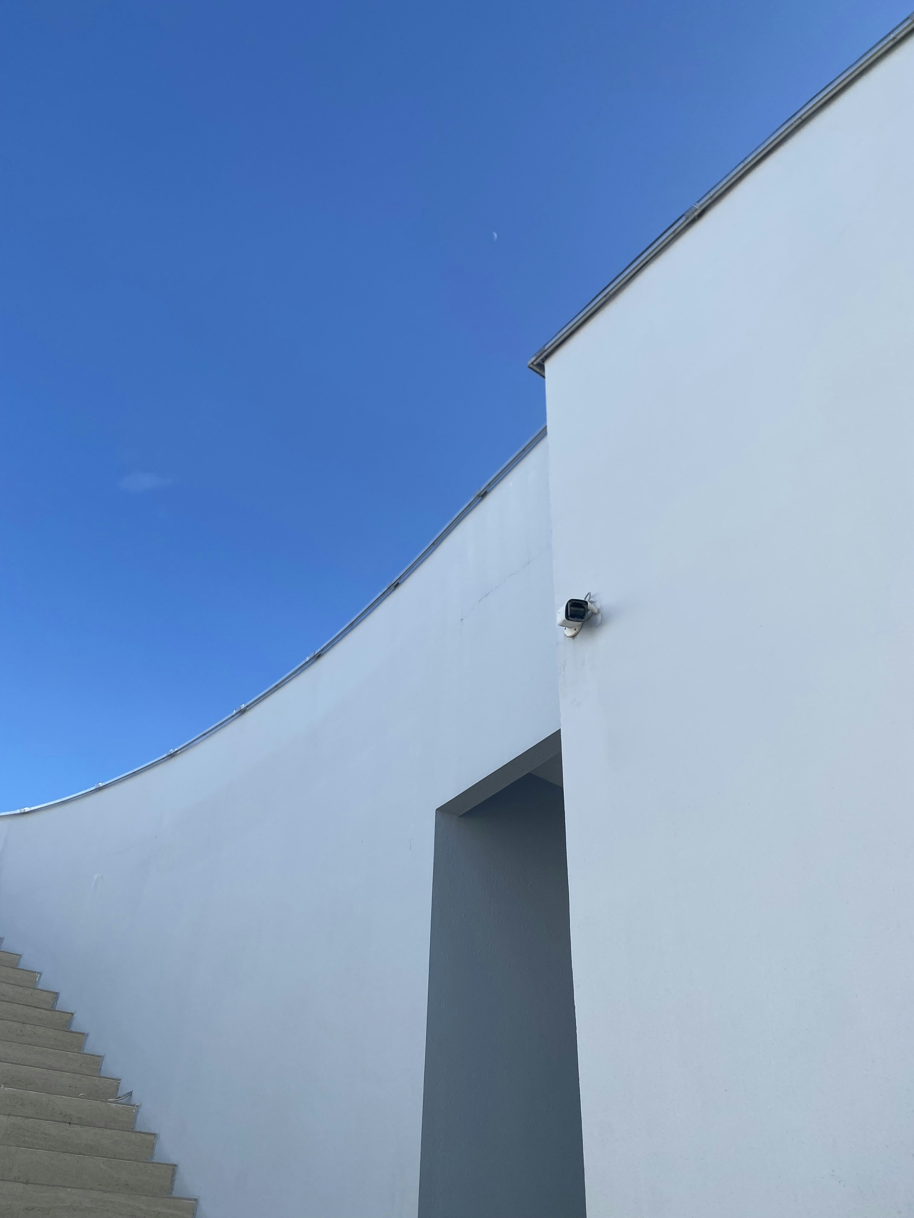 A minimalist architectural scene featuring a curved wall and a staircase leading to a doorway, set against a clear blue sky.