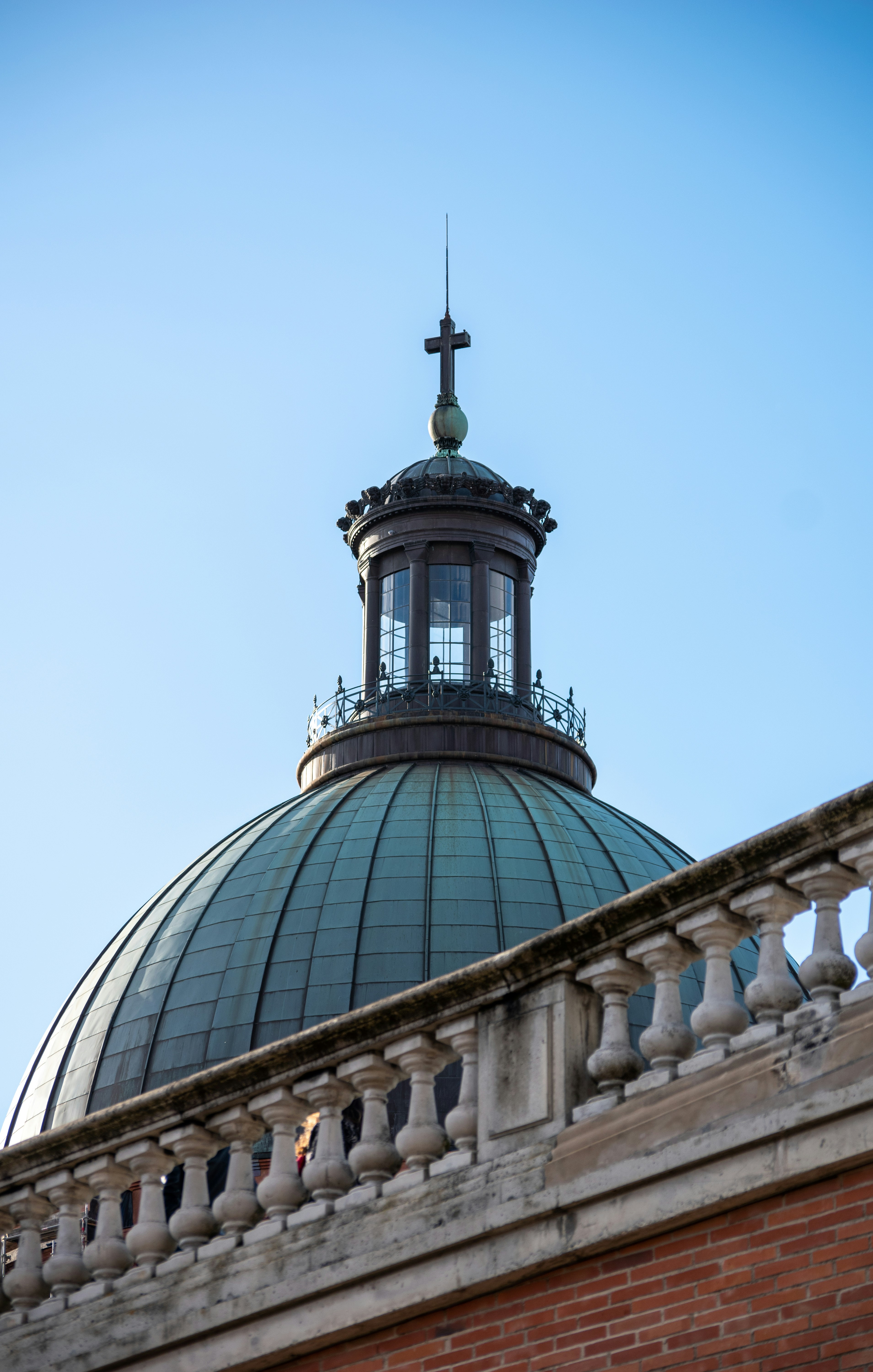 Copper-domed structure with a cross, framed by ornate stone balustrade under a clear blue sky.