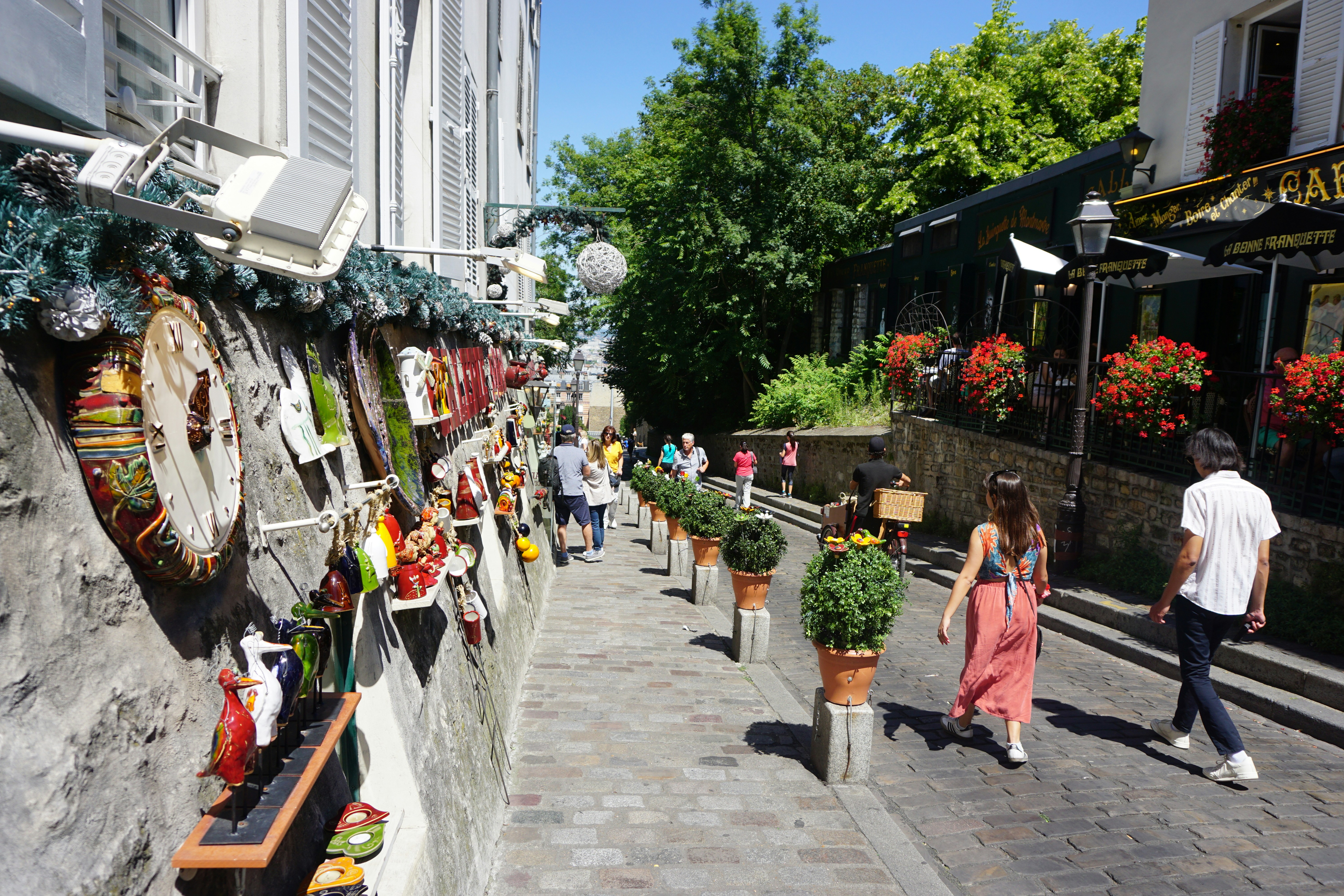 Cobblestone street with walls decorated with vibrant clocks and ceramics, flanked by potted plants and people walking.