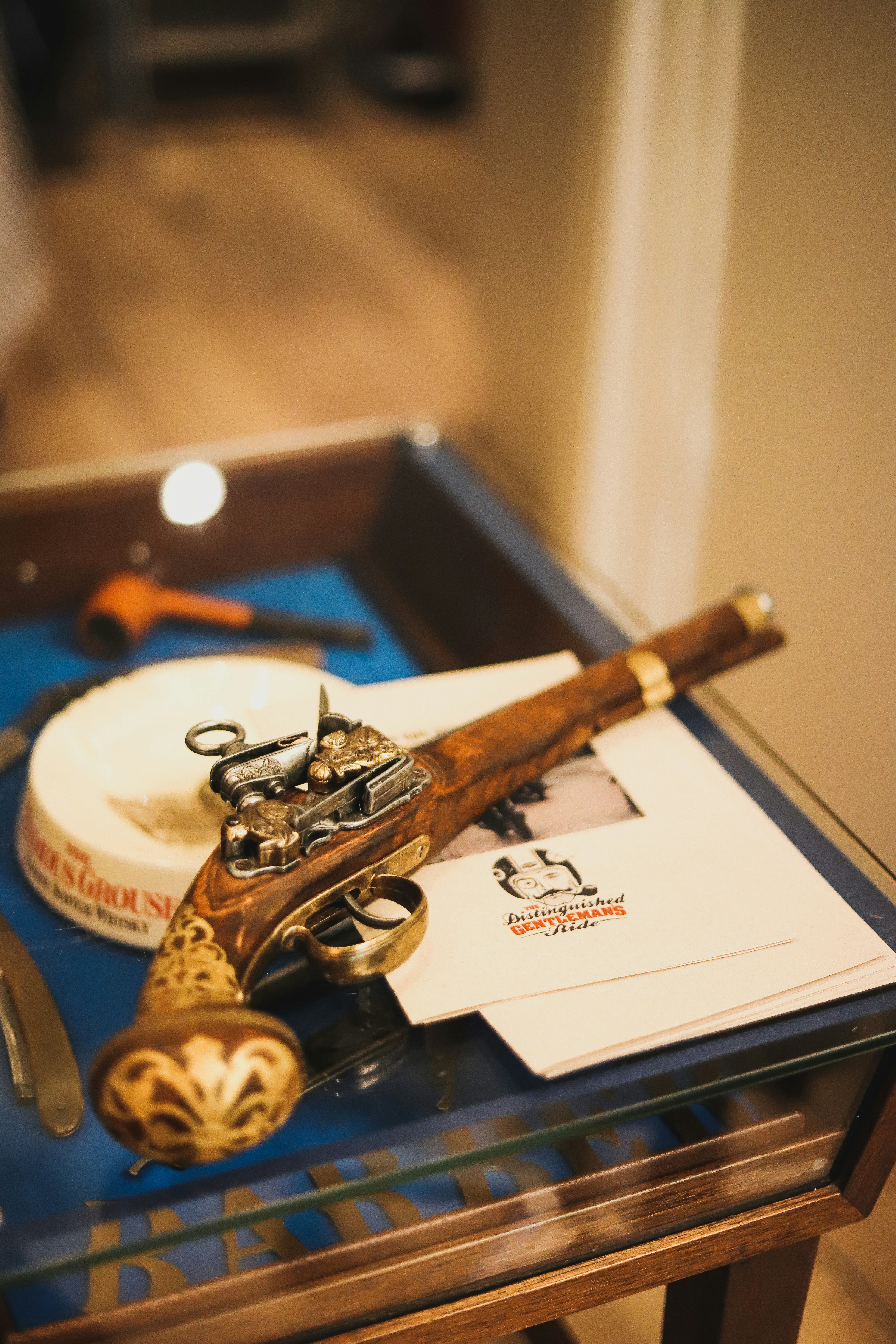 Close-up of an ornate flintlock resting on a stack of papers atop a glass-topped surface. A circular tin and a small tool sit nearby, adding contextual still-life details.