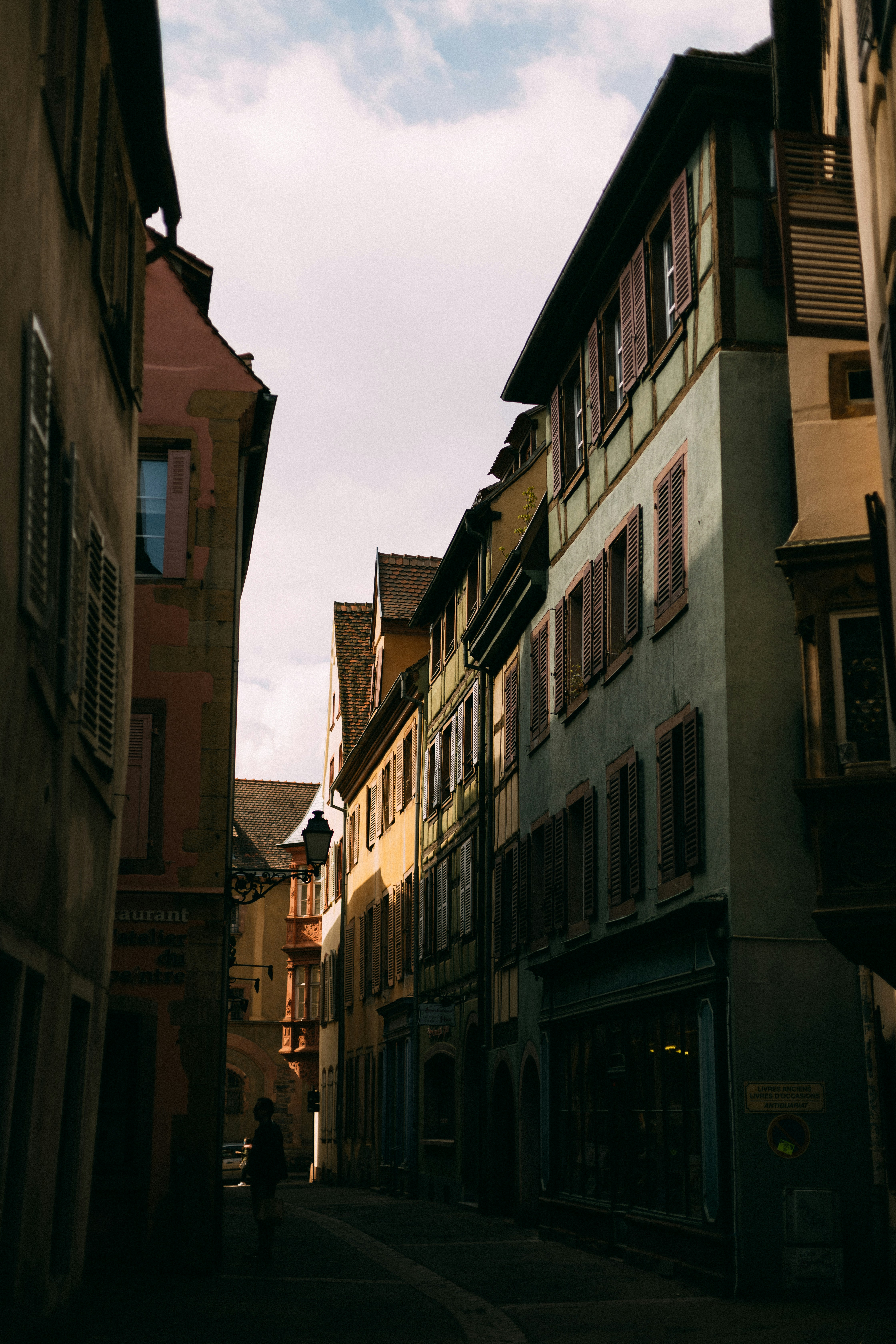 Charming narrow street flanked by colorful buildings with shutters, leading to a softly lit archway. A silhouette of a person adds a touch of life to the scene.