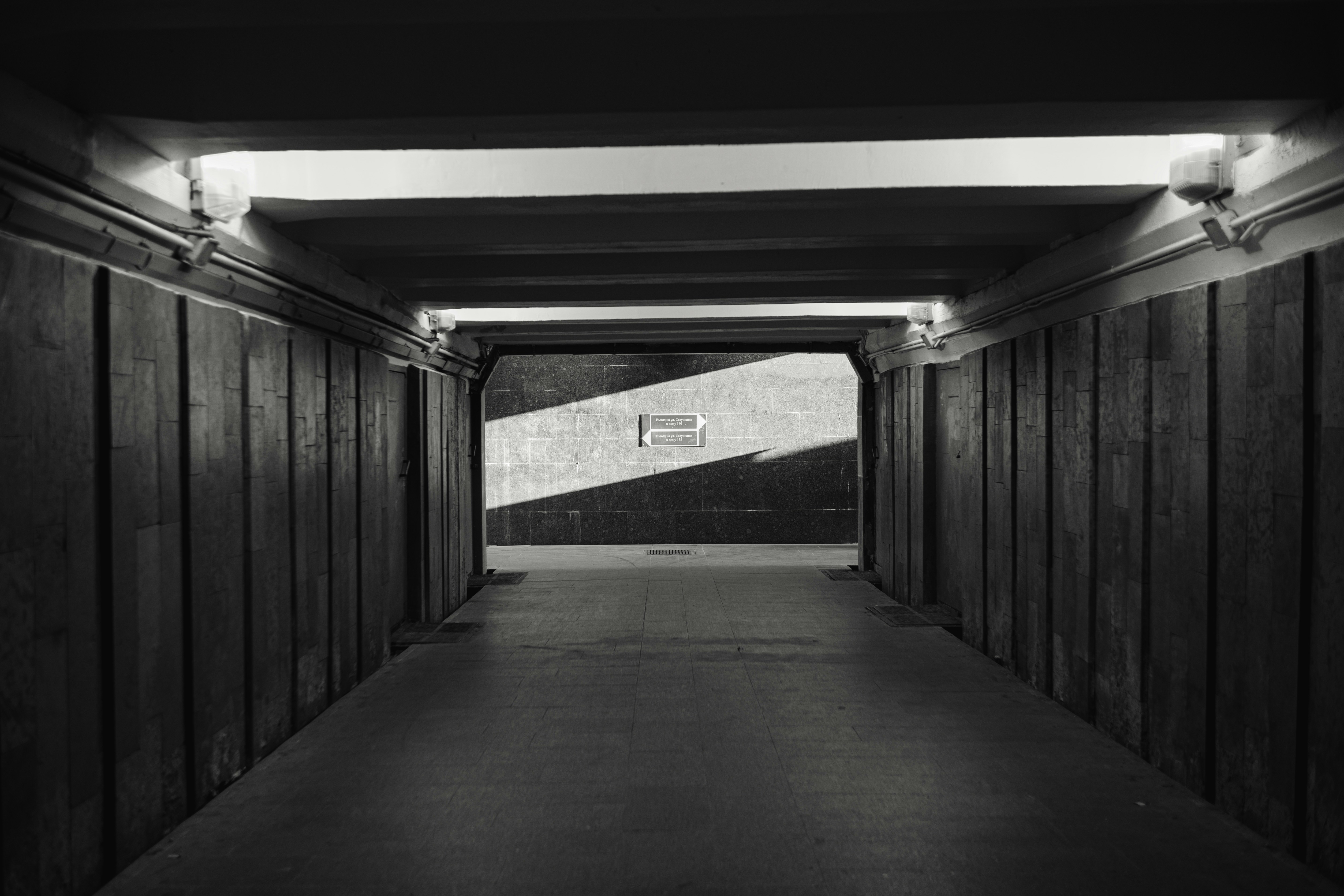 A dimly lit corridor with geometric patterns on the walls, leading to a starkly illuminated area at the end. The contrast between light and shadow creates a dramatic atmosphere.