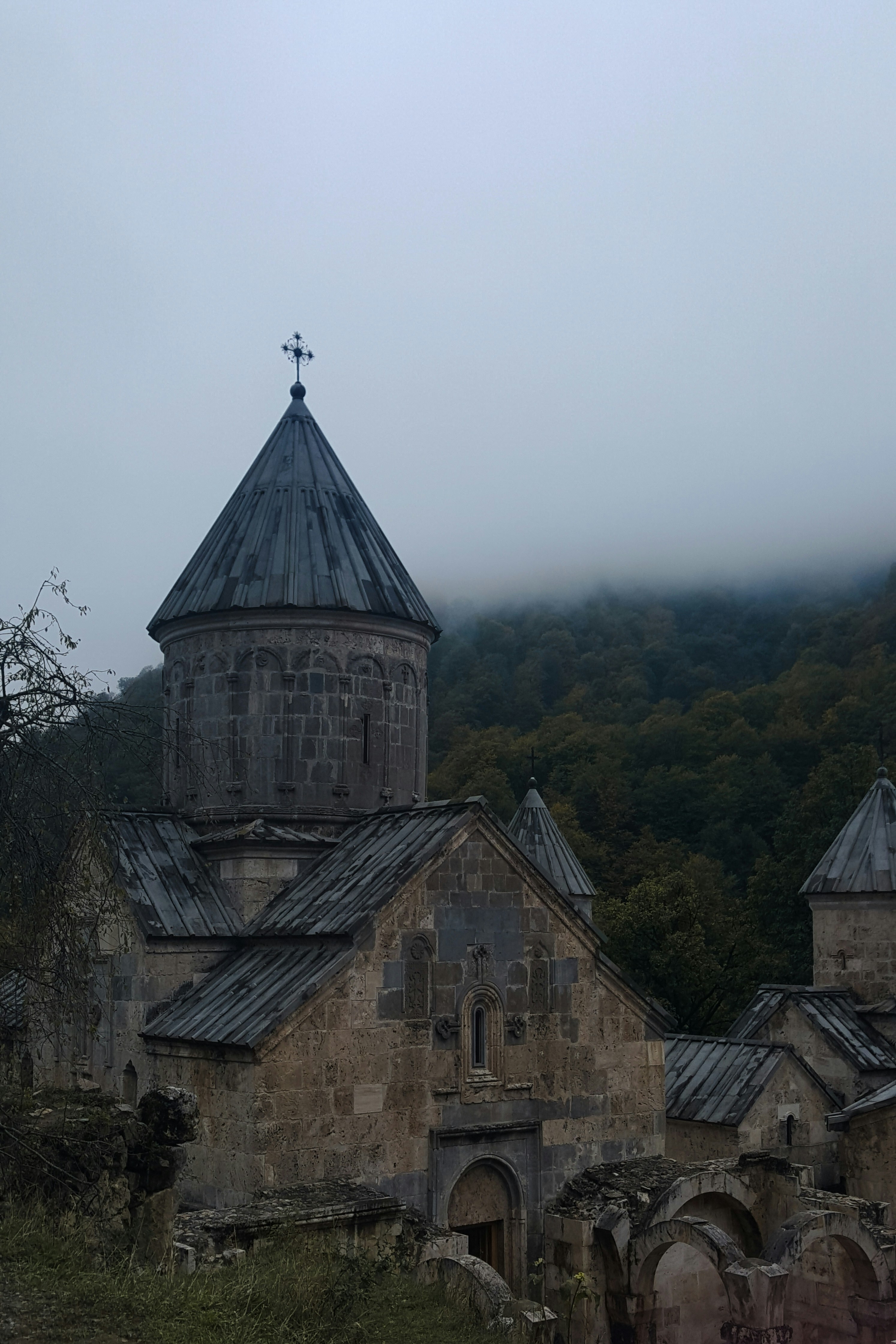 Ancient stone church with a conical turret rises against fog-enshrouded hills. The image highlights weathered masonry, textured roofing, and a moody, atmospheric scene.
