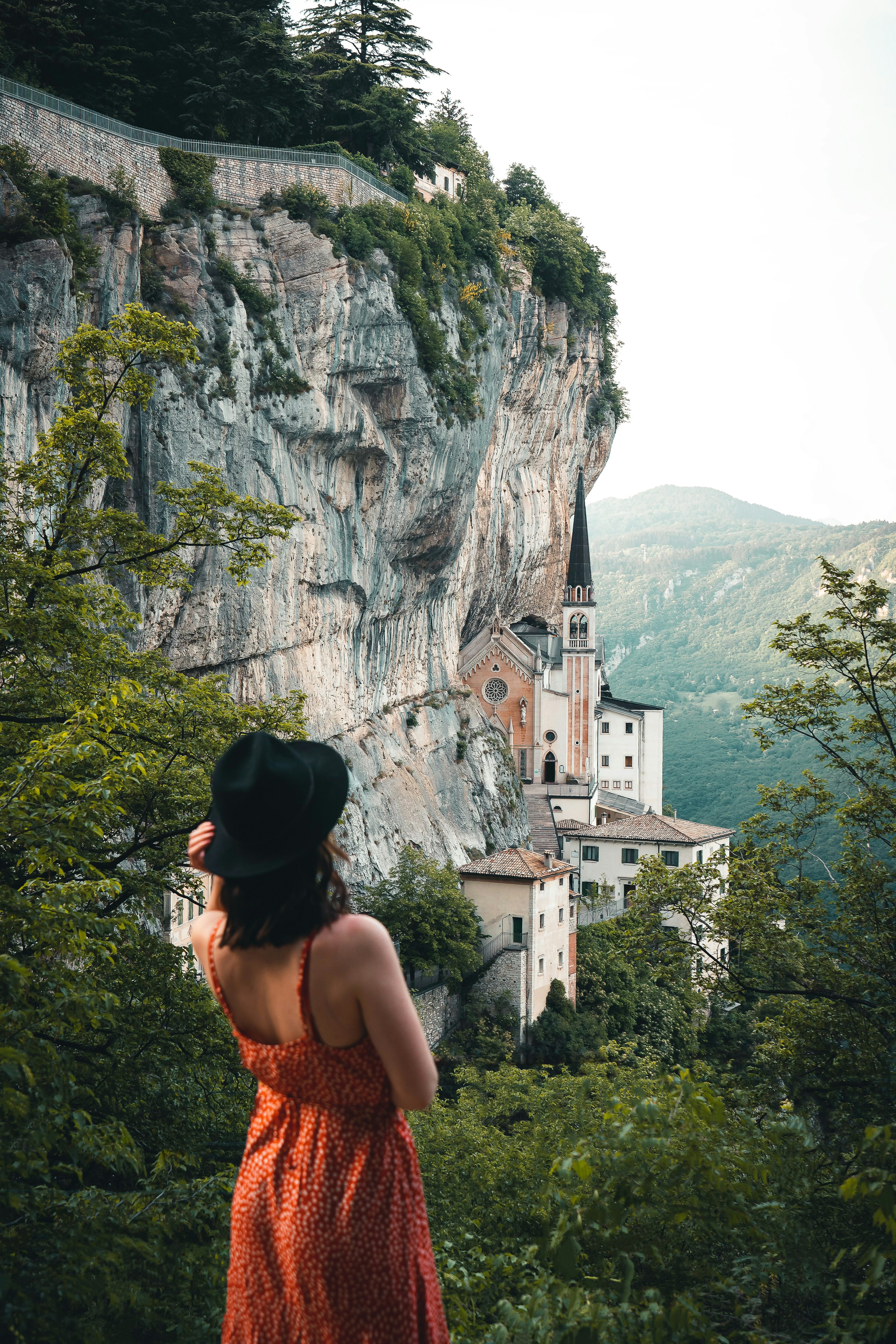 A woman in a red dress gazes at a stunning church nestled against a rocky cliff, surrounded by lush greenery and mountains in the background.
