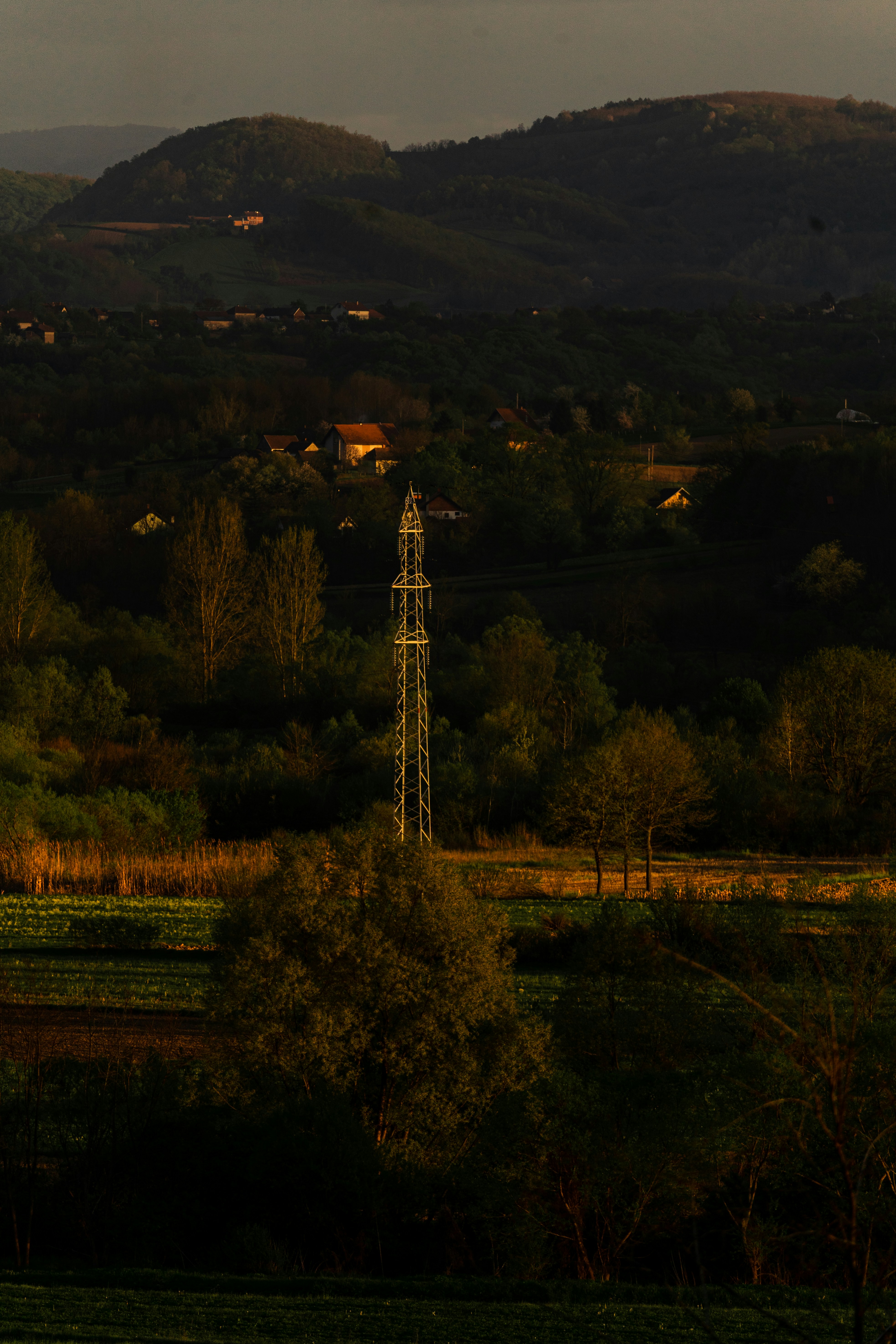 A solitary powerline tower rises amidst rolling hills and lush greenery, capturing the serene essence of rural life at dusk.