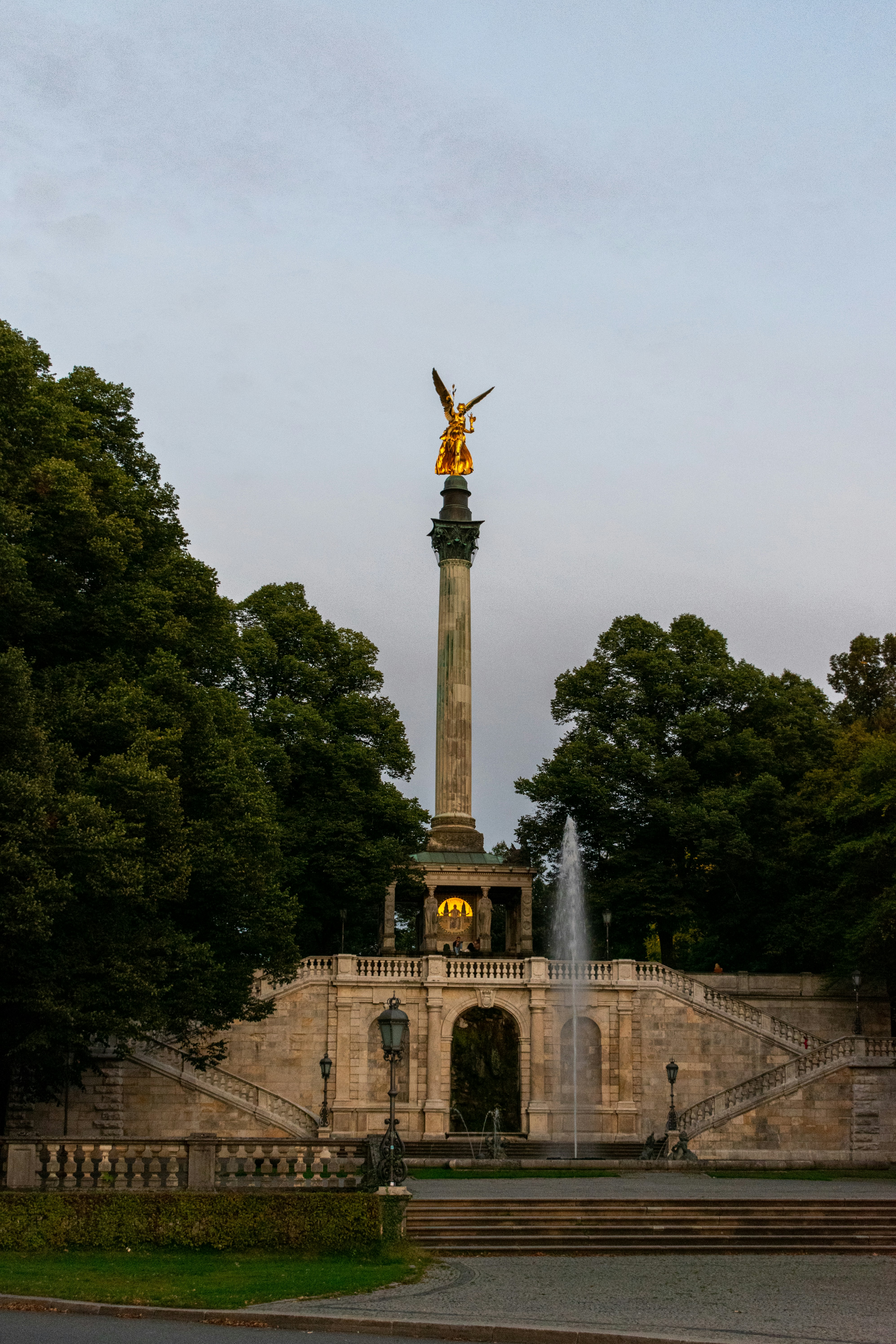 A majestic statue of an angel atop a tall column, surrounded by lush greenery and a cascading fountain, symbolizing victory and peace.